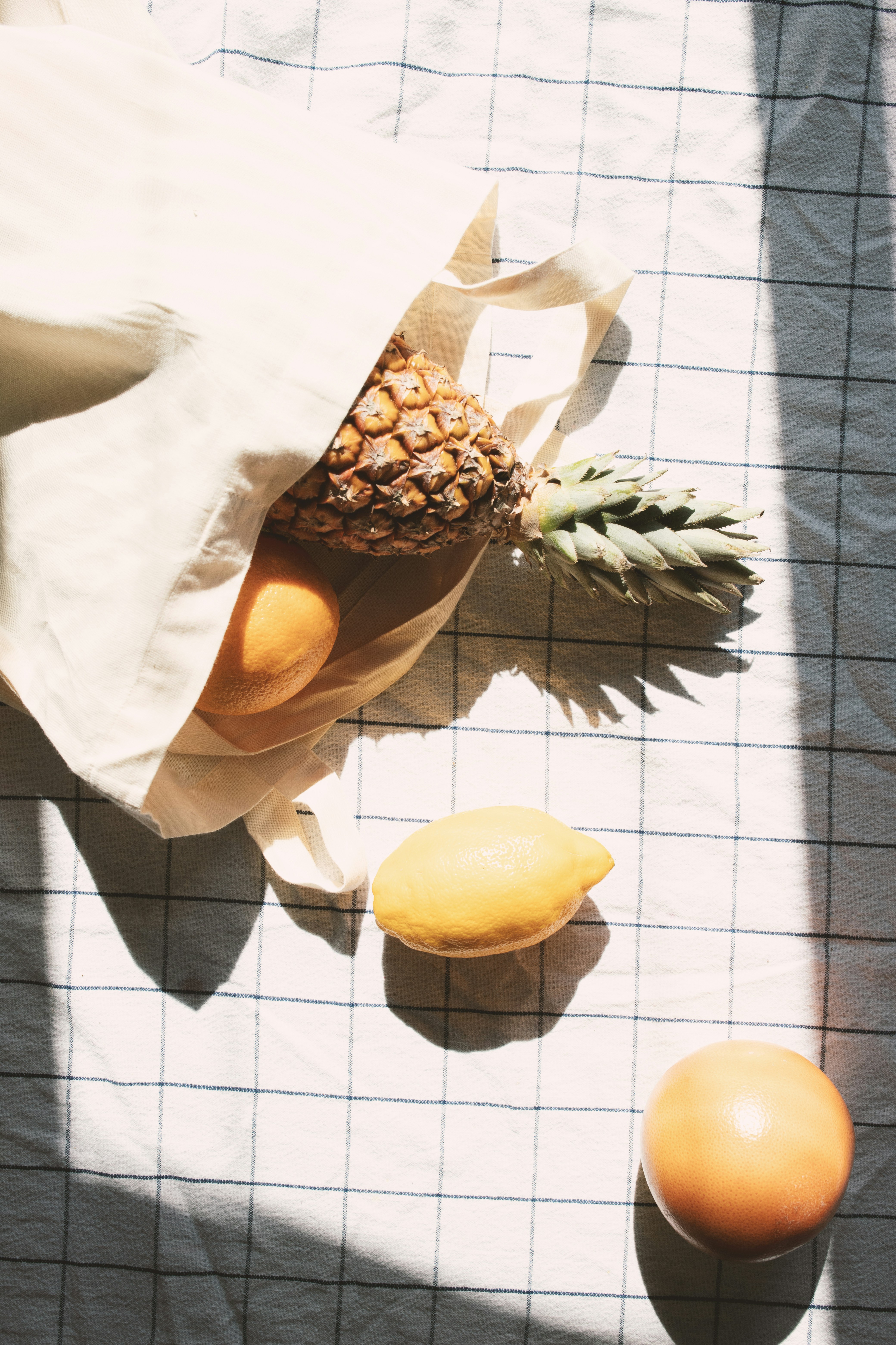 Sunlit Harvest: Pineapple, Mango, and Citrus spill from a tote on a checkered cloth