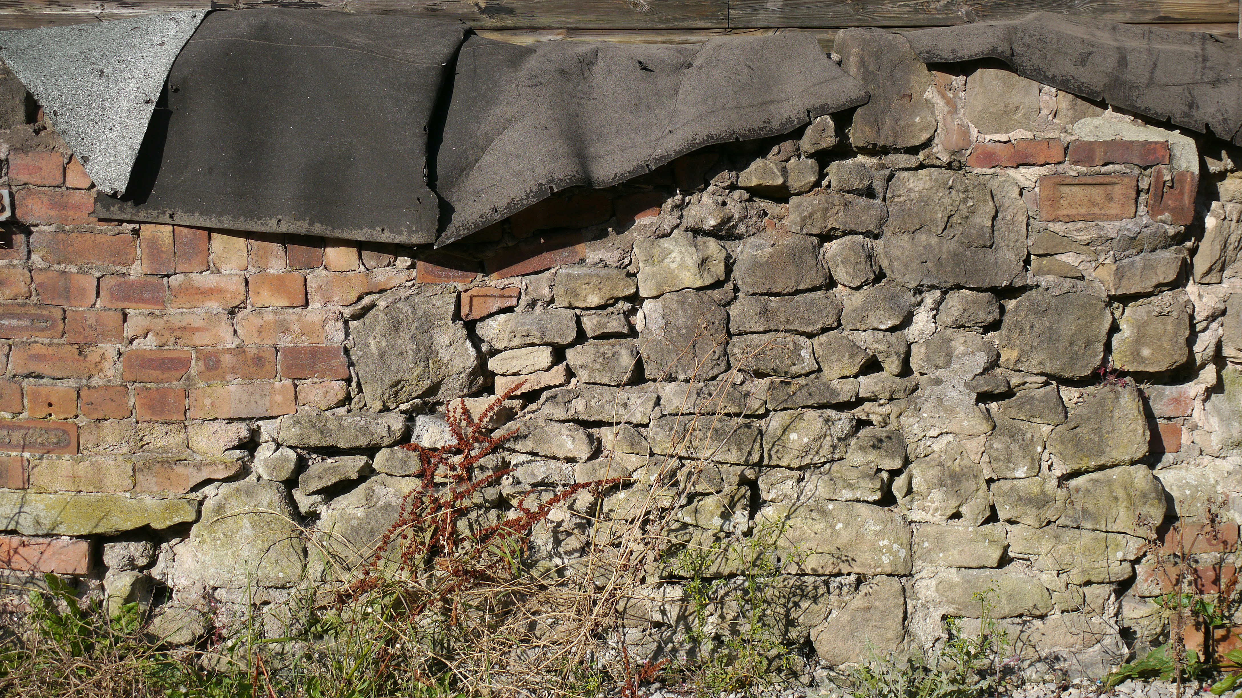 A textured wall showcasing a mix of aged bricks and stones, partially covered by a tattered tarp, with wild grasses peeking through the cracks.