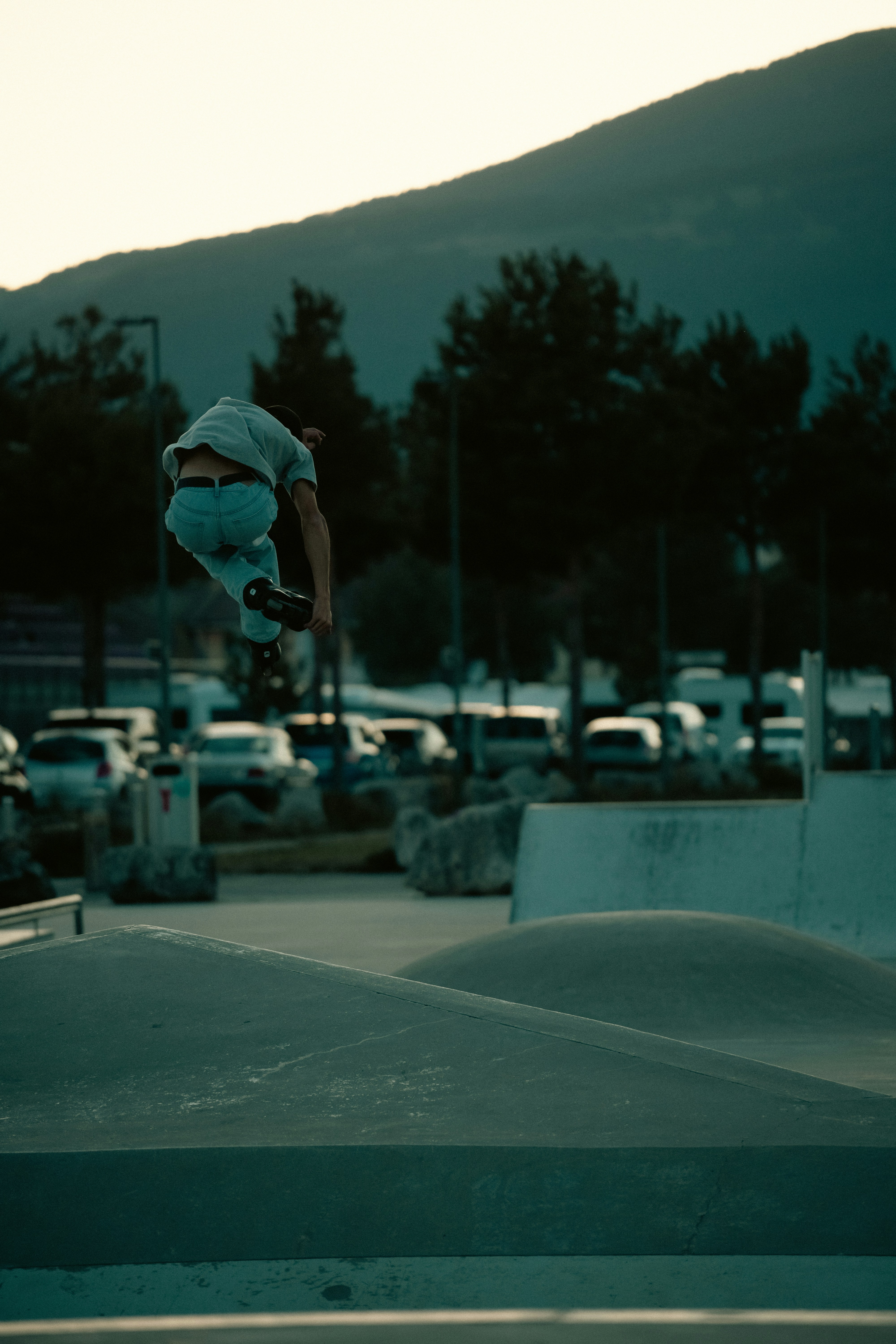 Skateboarder mid-air against a backdrop of mountains and trees at dusk.