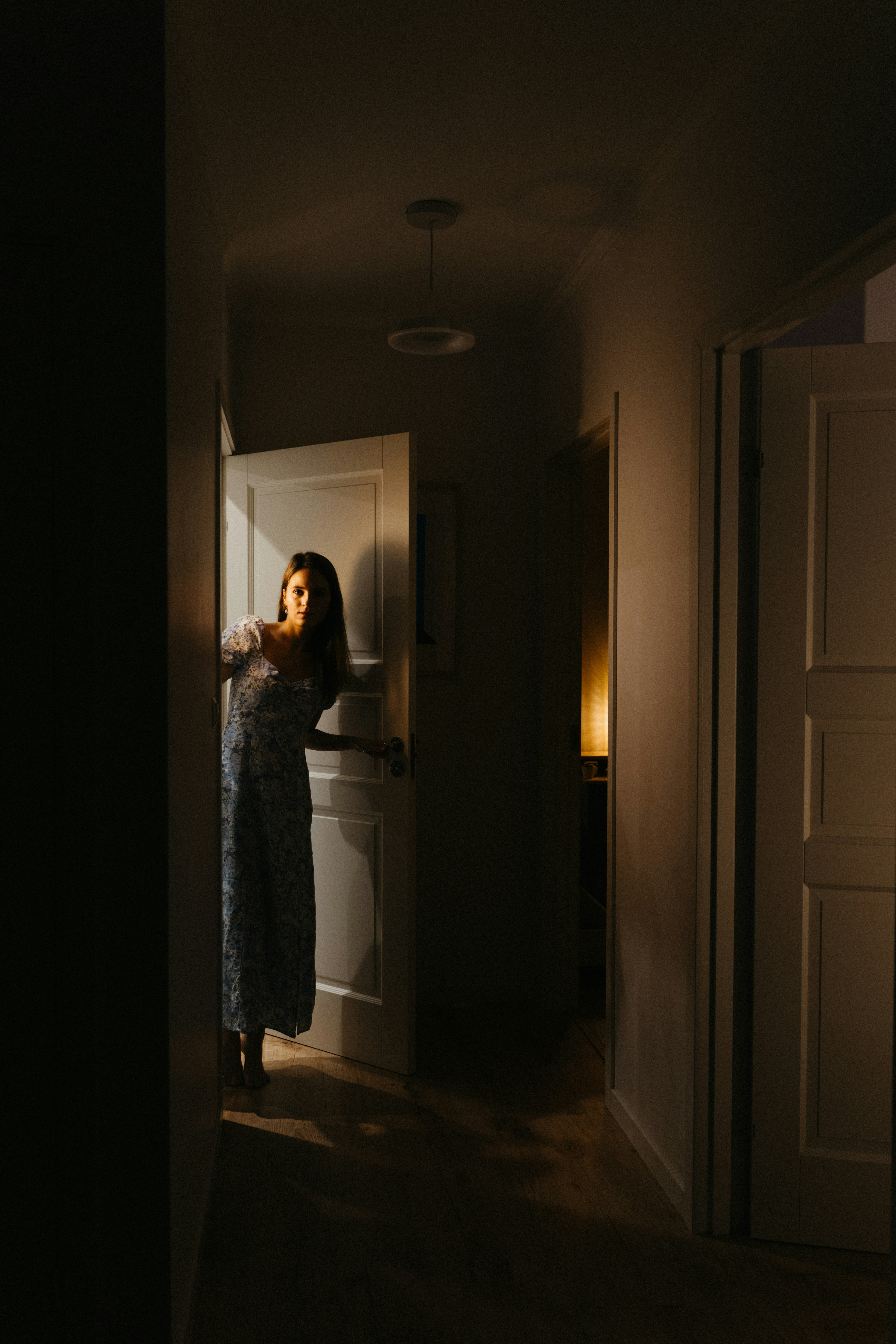 A woman in a floral dress peeks through an open door, illuminated by soft light in a dim hallway. The contrasting shadows create a sense of intrigue.