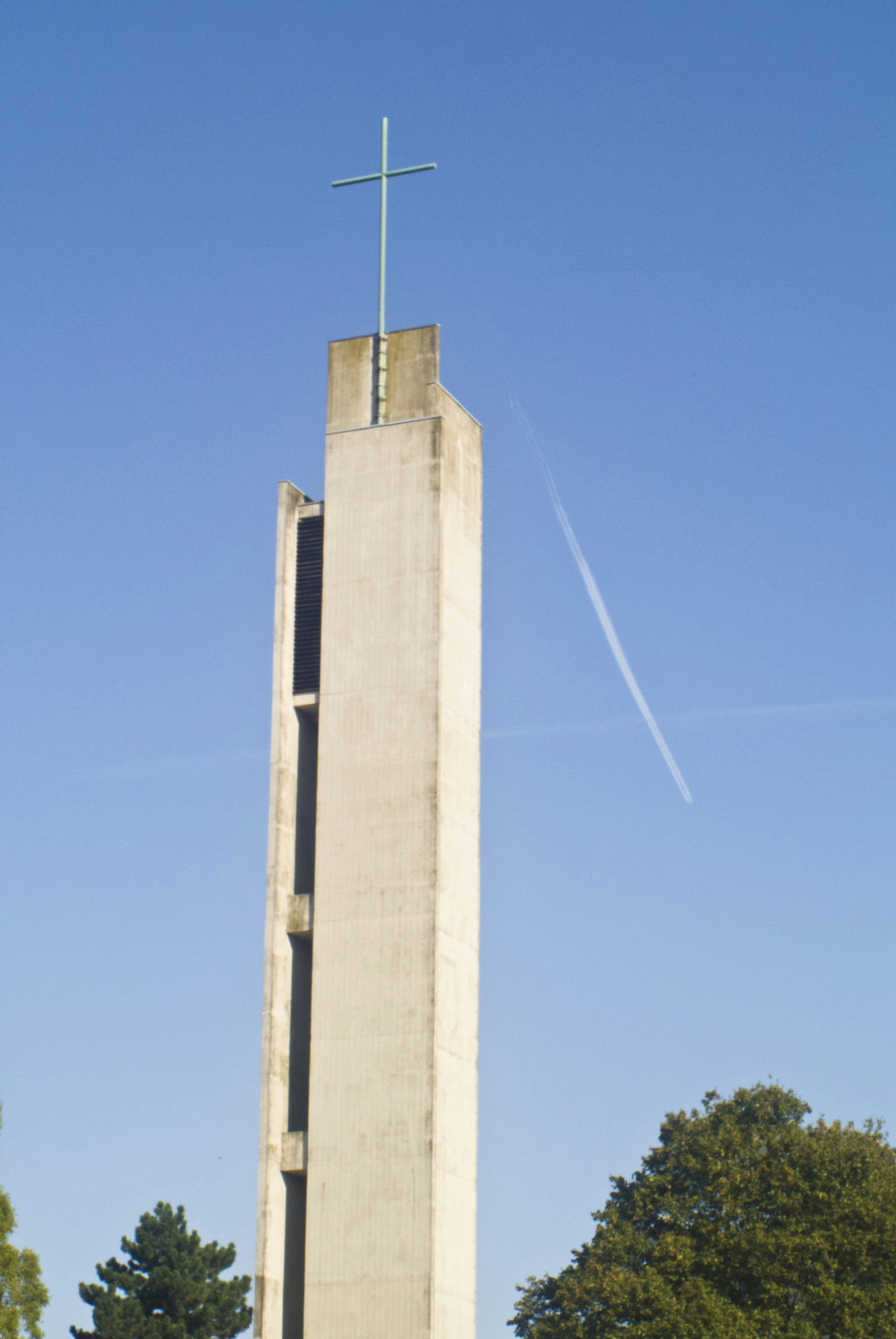 Tall concrete church tower with a cross on top rises against a clear blue sky, framed by trees.