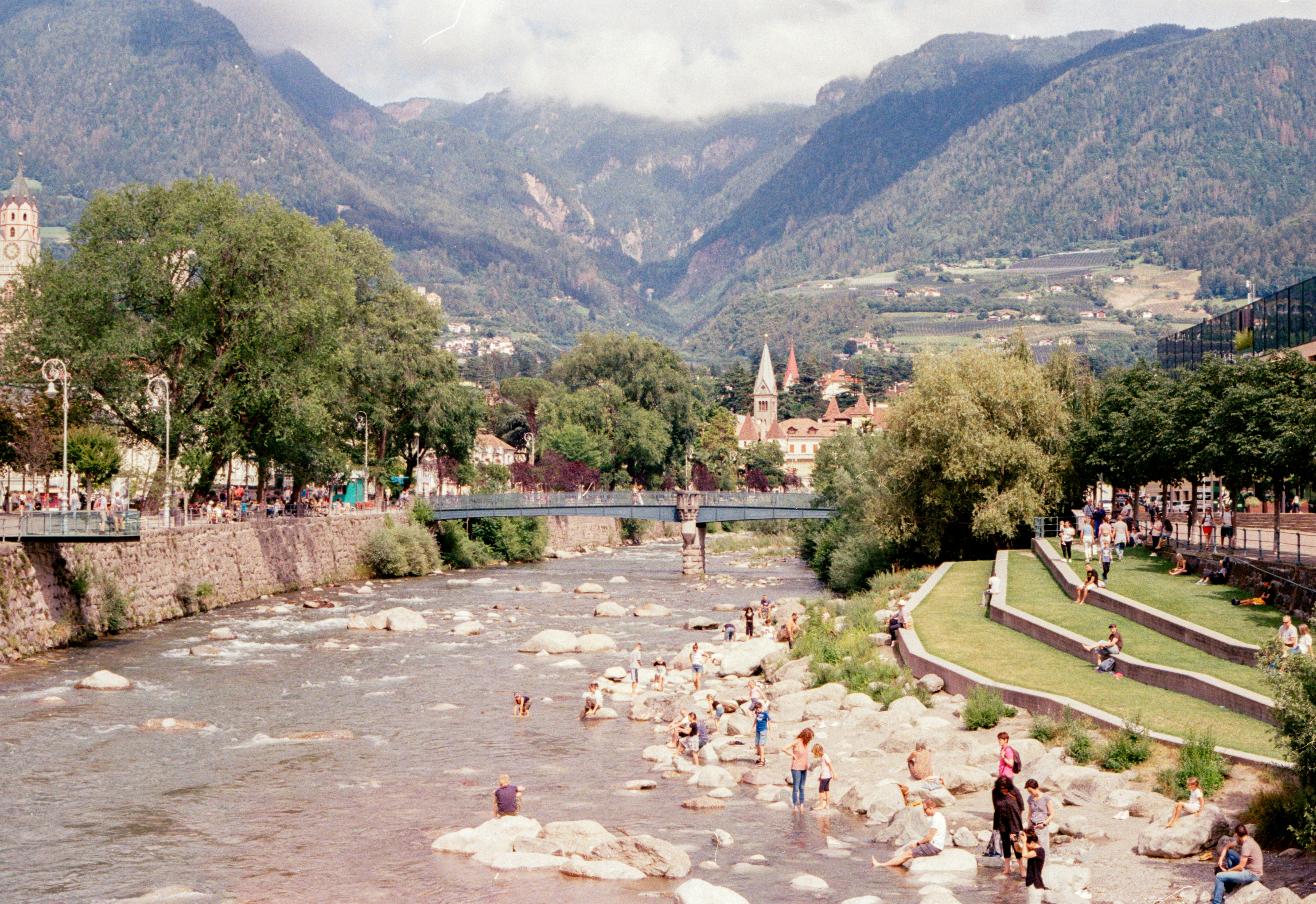 Families and friends enjoying a sunny day by the river, surrounded by lush greenery and distant mountains. The scene captures a harmonious blend of nature and leisure activities.