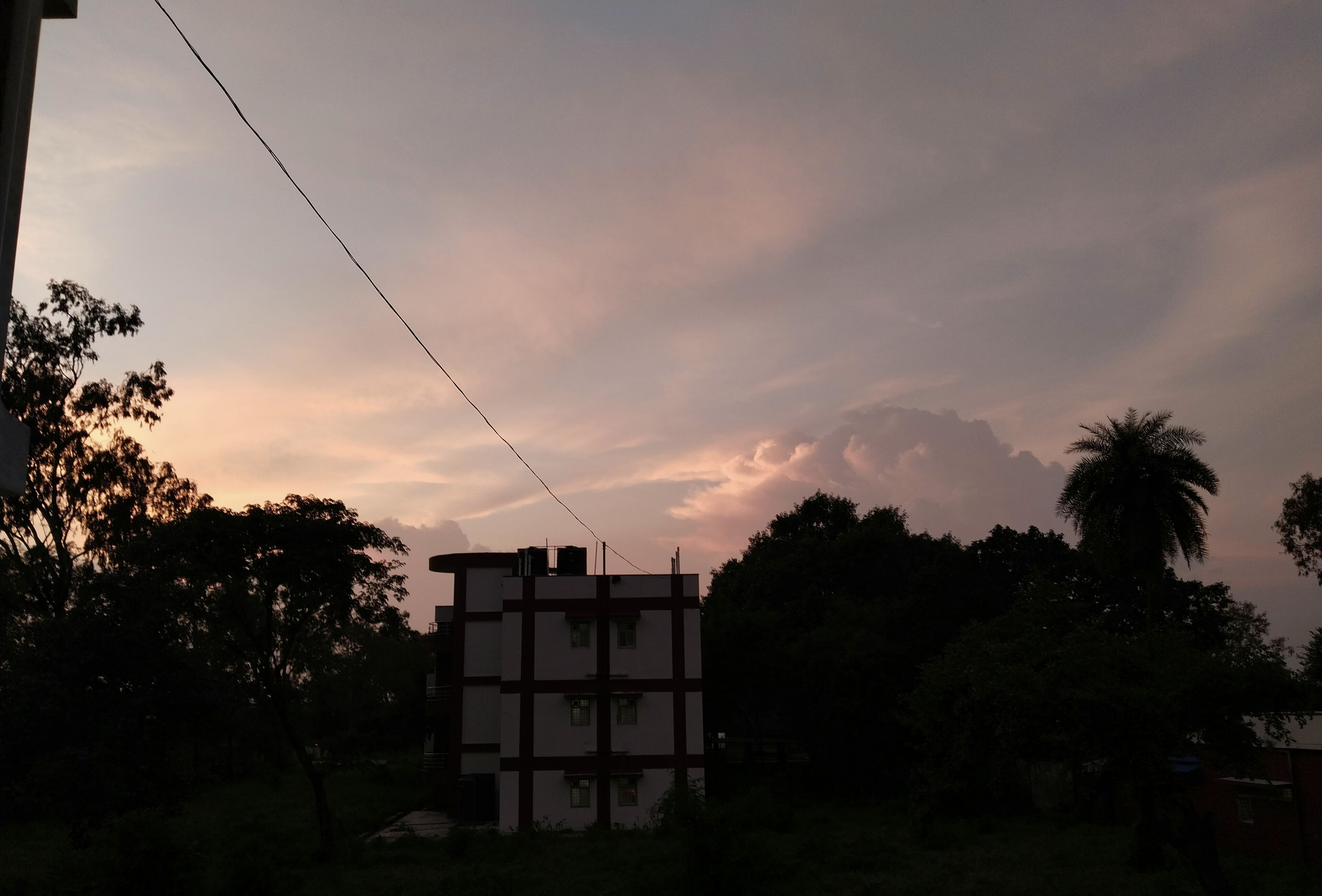 Silhouetted trees frame a small, blocky building against a pastel sunset, creating a calm rural horizon. The scene emphasizes dusk mood and soft lighting.