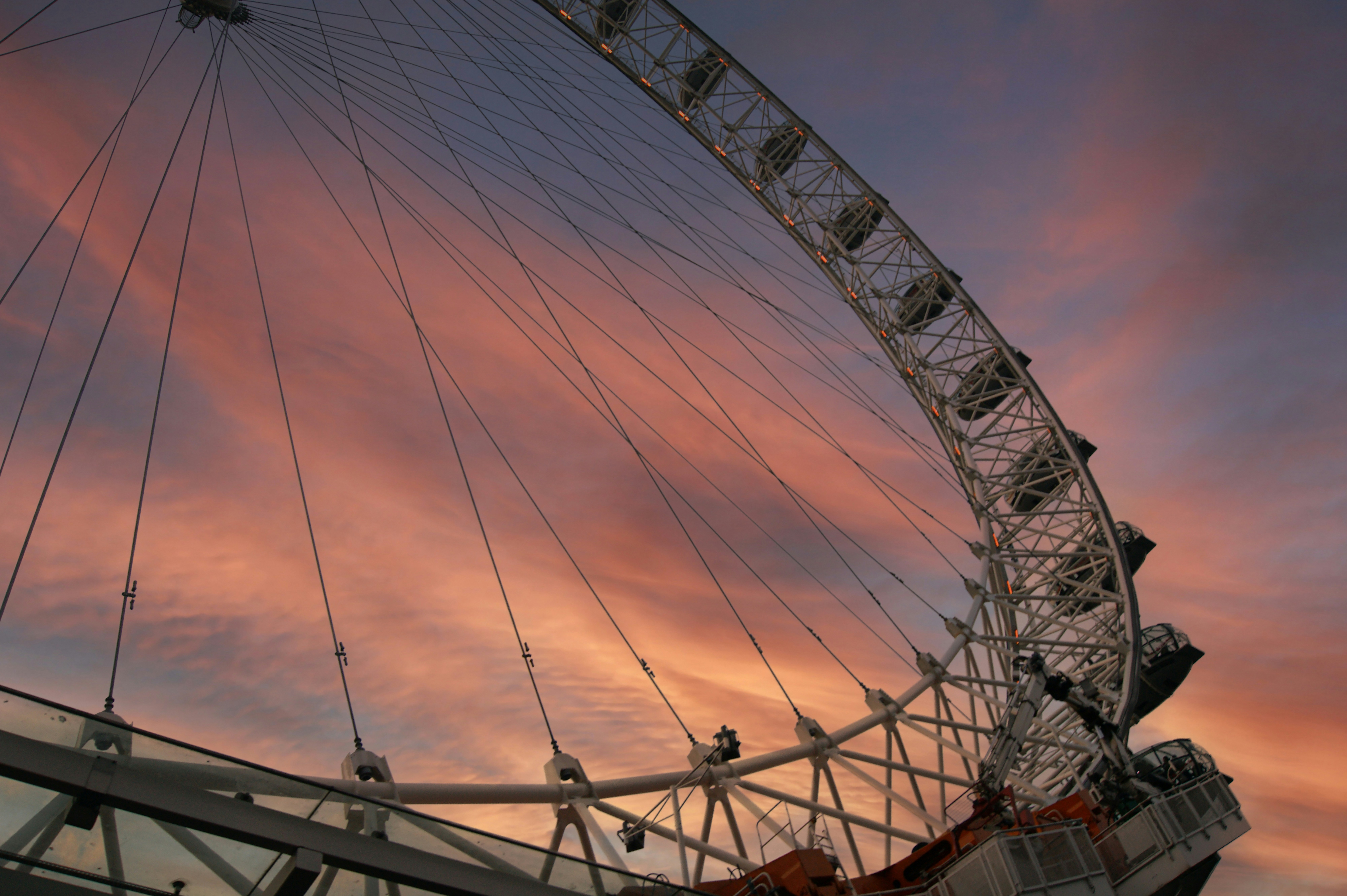 Ferris wheel arc dominates the frame, its spokes radiating toward a warm orange-pink sky. The silhouetted metal structure emphasizes geometric lines against the glowing horizon.