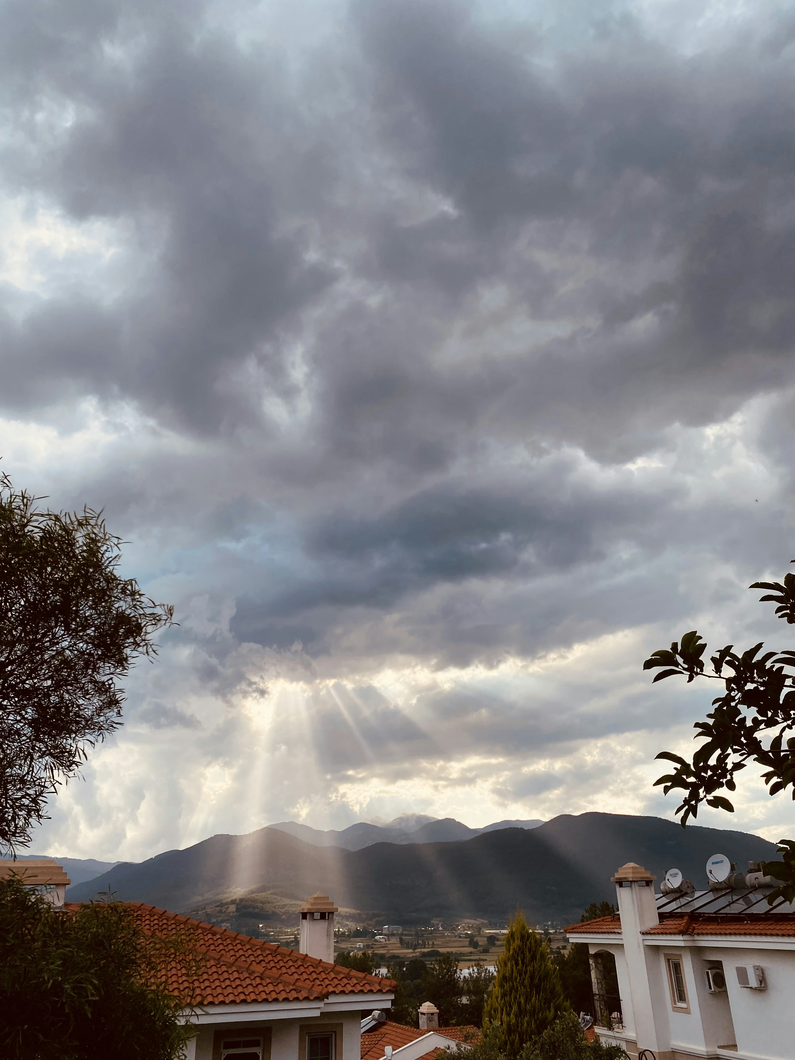 Sunbeams breaking through dramatic clouds illuminate the mountain range, creating a serene atmosphere. The scene captures the tranquil beauty of nature at dusk.