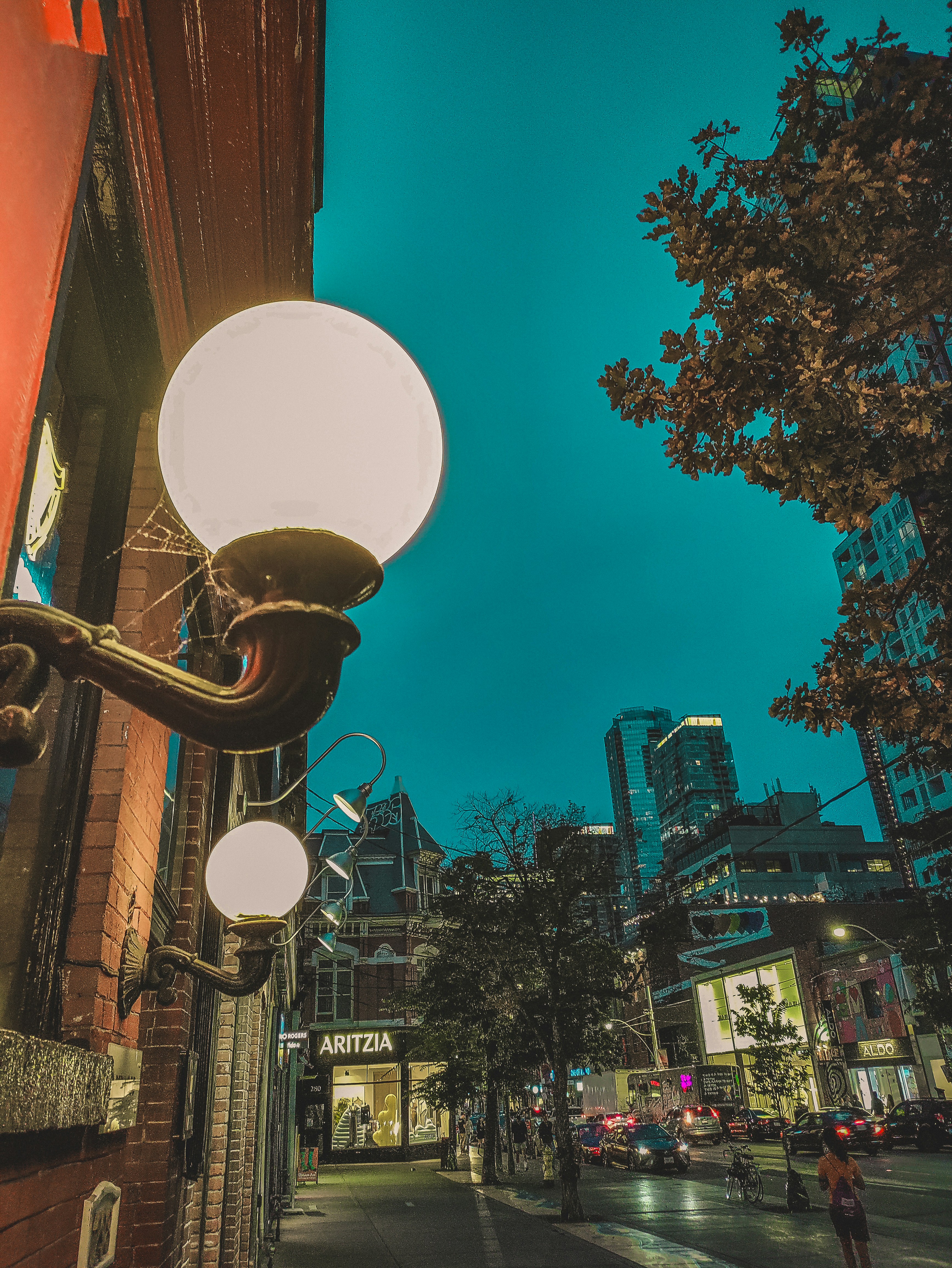 Nighttime city street photograph with warm globe lamps along a brick facade, leading the eye toward modern towers and storefronts.