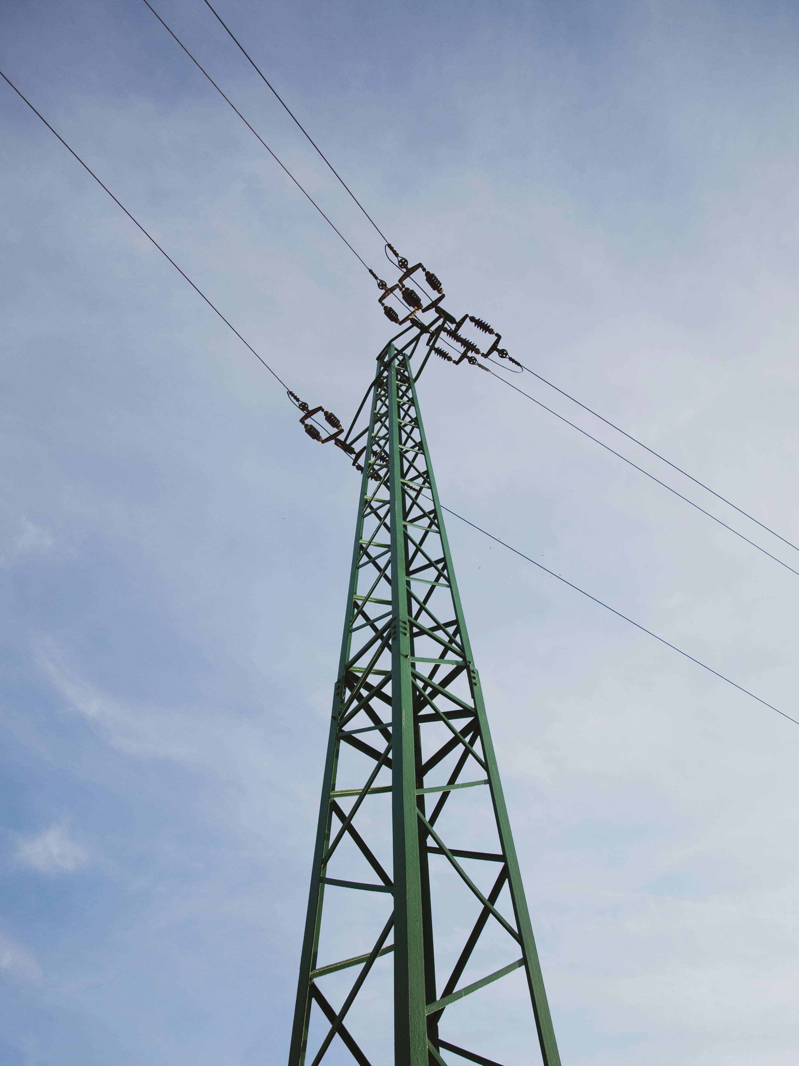A photograph of a green lattice transmission tower reaching toward a clear blue sky, with power lines crossing diagonally across the frame.