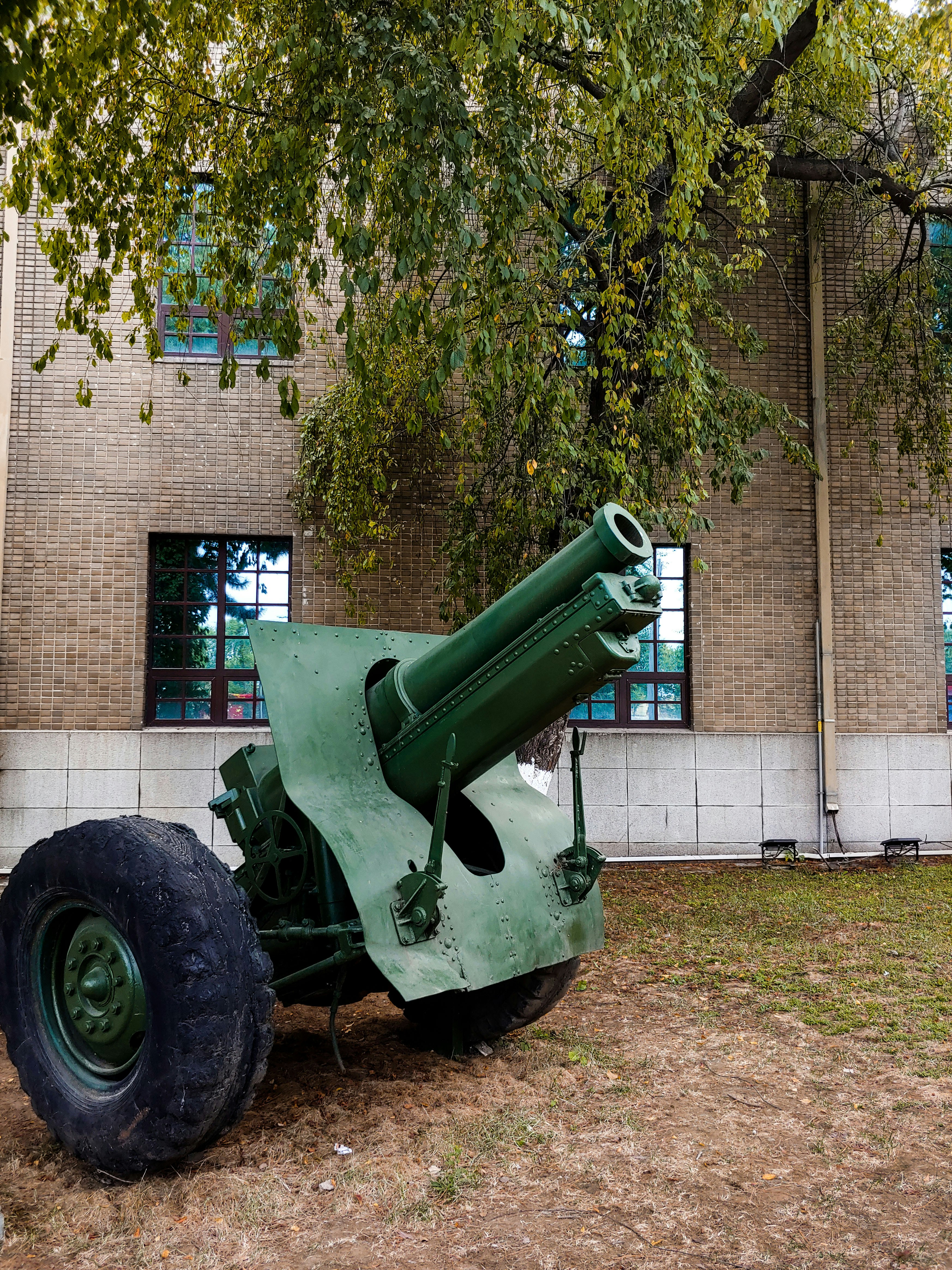 Green artillery cannon leaning on its wheel in front of a brick building with windows, under leafy trees; a static, historical exhibit outdoors.