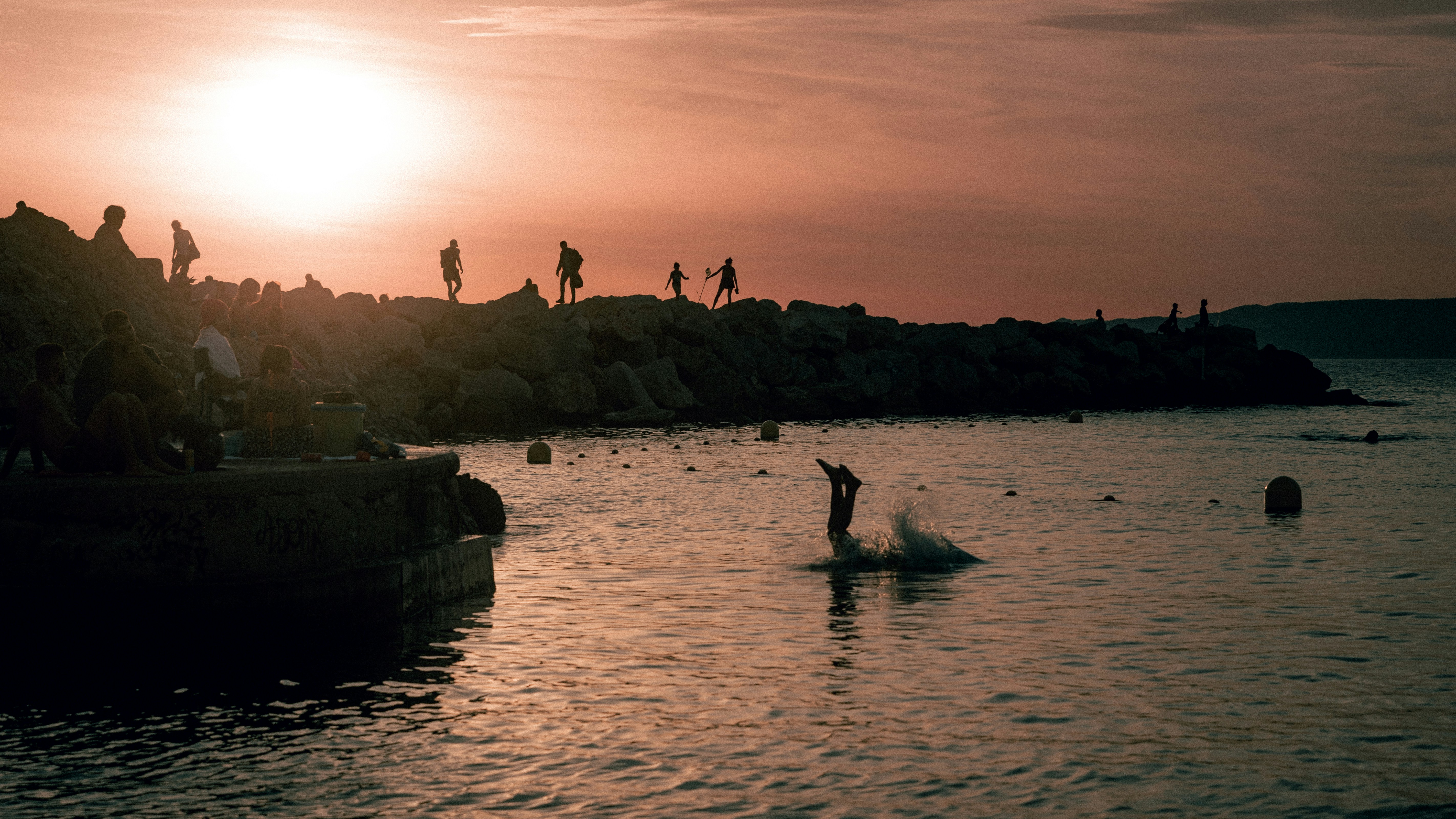 Silhouette of a person diving into calm waters at sunset, with a rocky shoreline and distant figures.