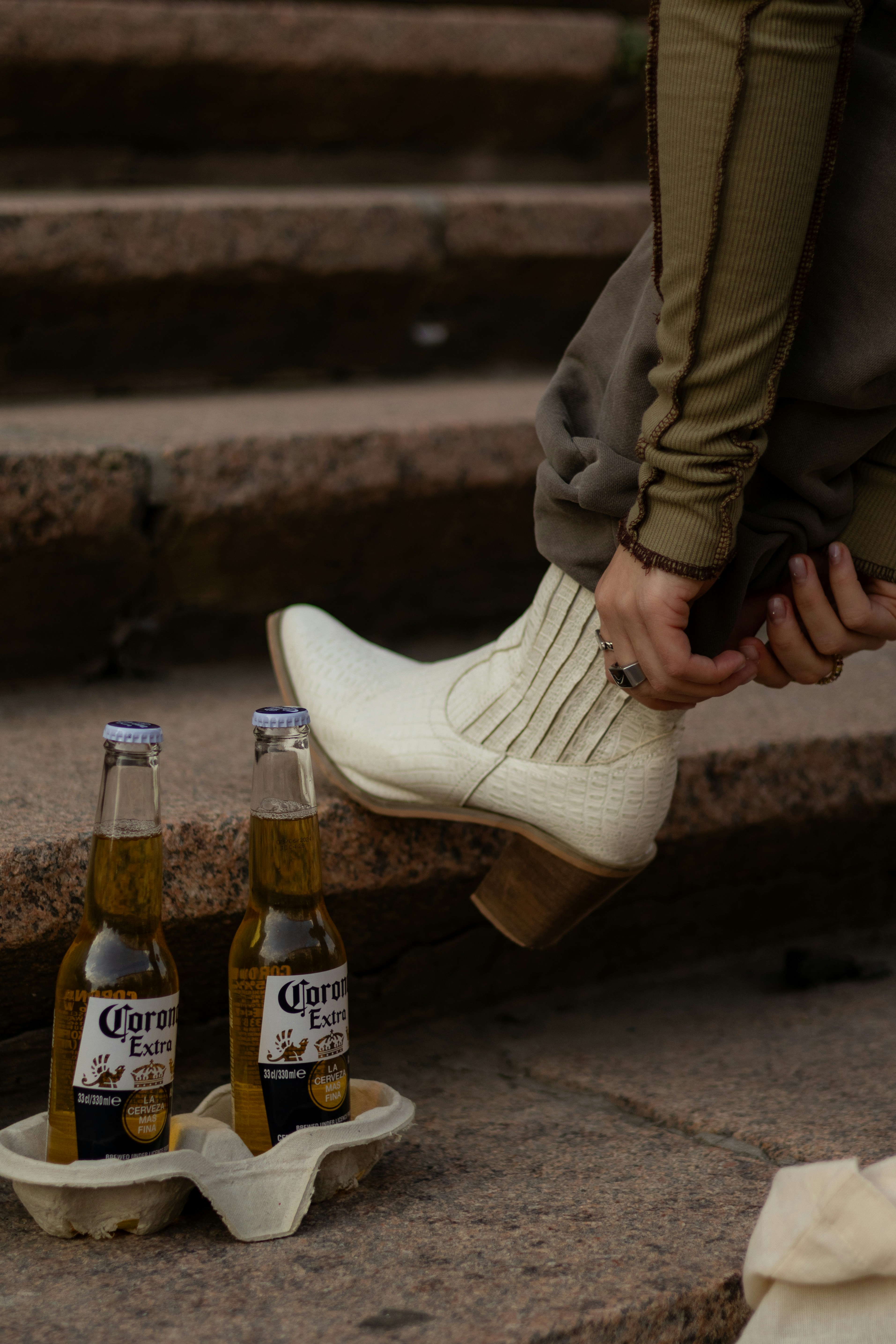 A person ties a white ankle boot on weathered stone steps while two Corona bottles sit in a carton nearby.