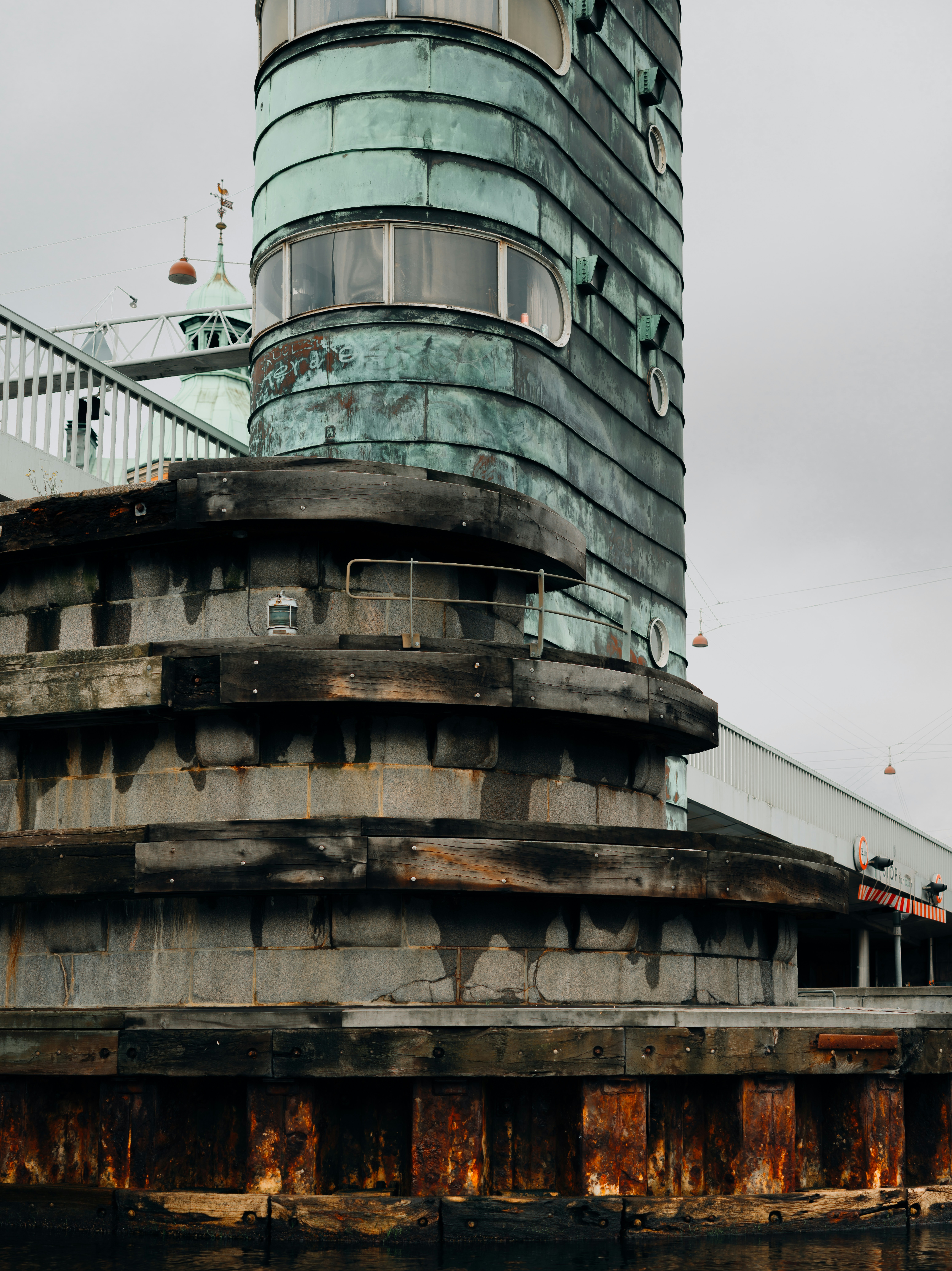 Weathered verdigris turret rising from a rusted harbor pier with stacked circular decks. The scene emphasizes industrial decay against a muted sky.