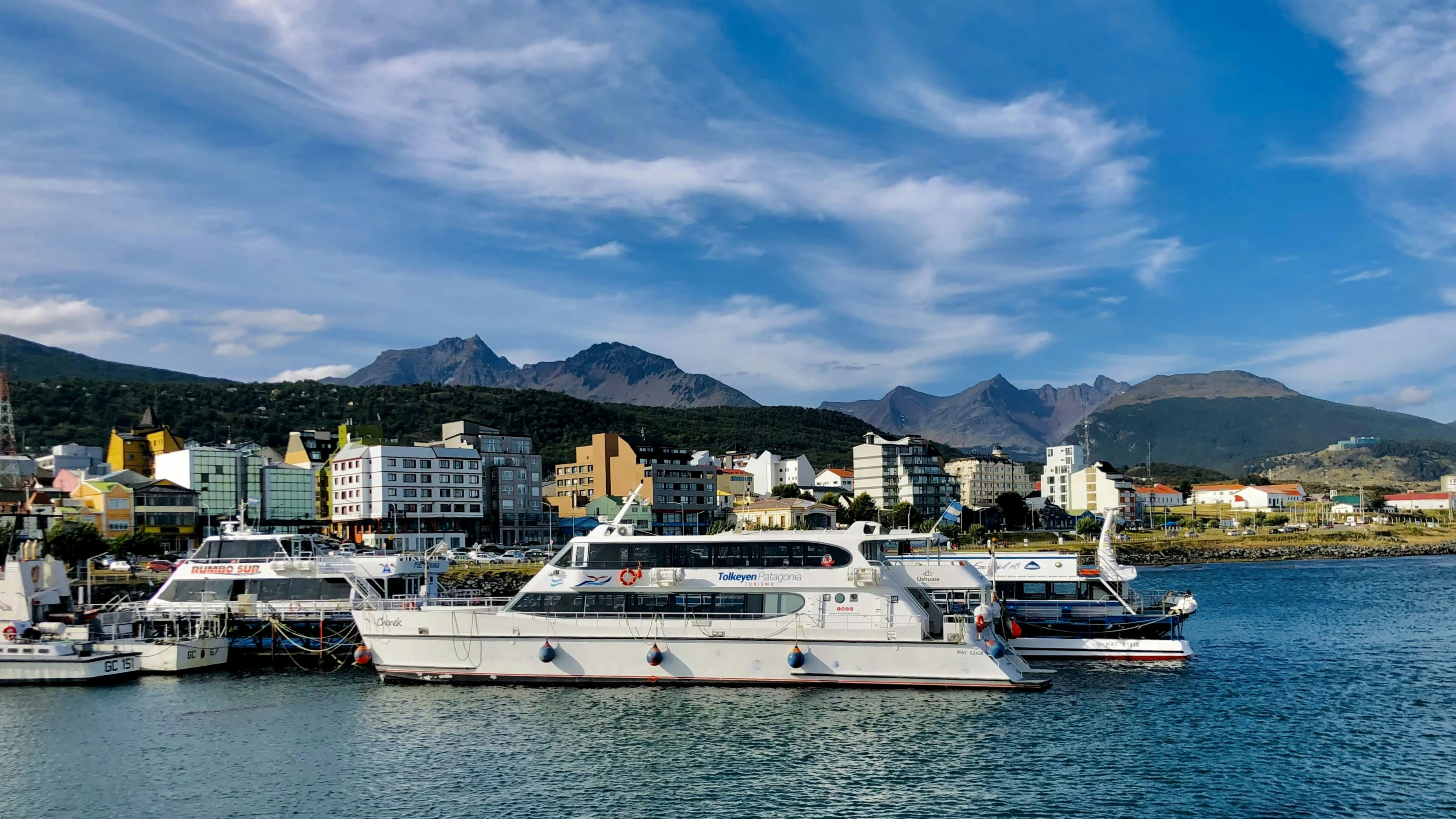White yachts docked in a tranquil harbor with a backdrop of city buildings and distant mountains under a vivid blue sky.