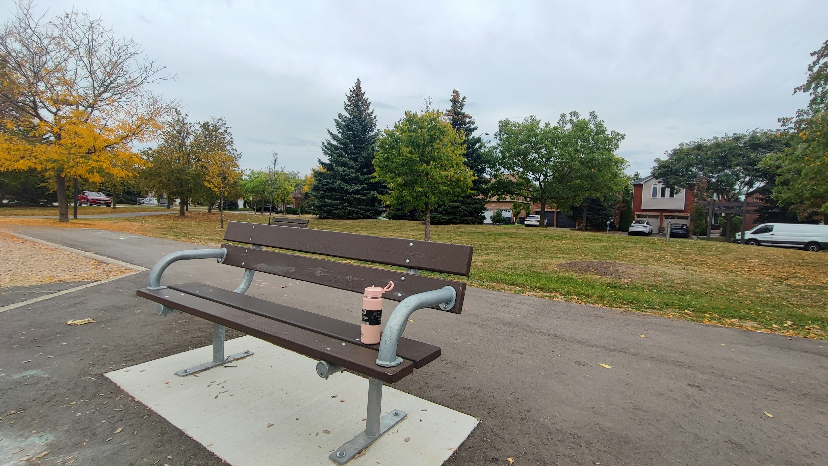 A solitary park bench on a concrete pad with a pink water bottle rests under autumn trees in an overcast park.