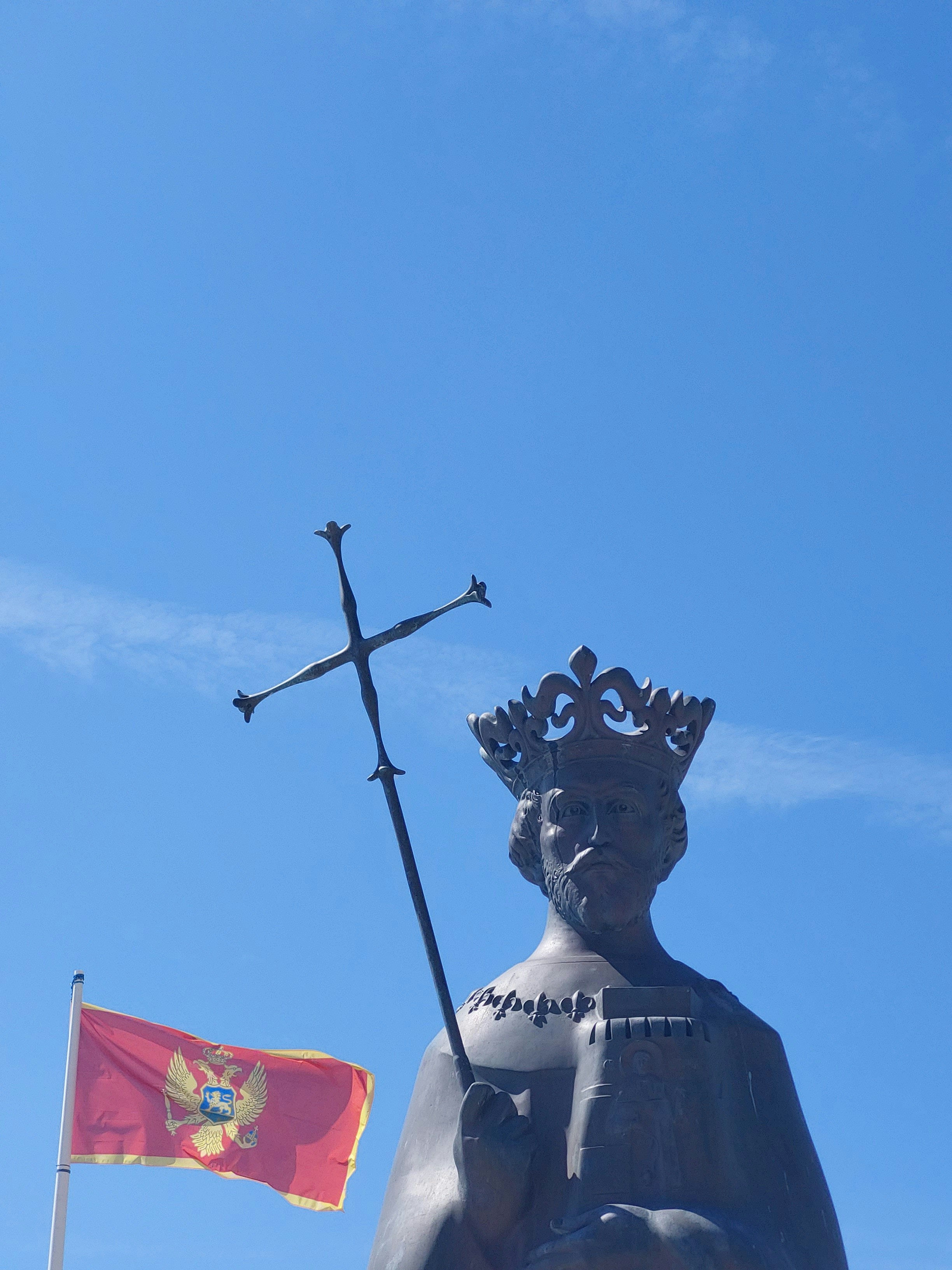 Bronze statue of a crowned king holding a tall cross, set against a bright blue sky with a Montenegro flag in the foreground.