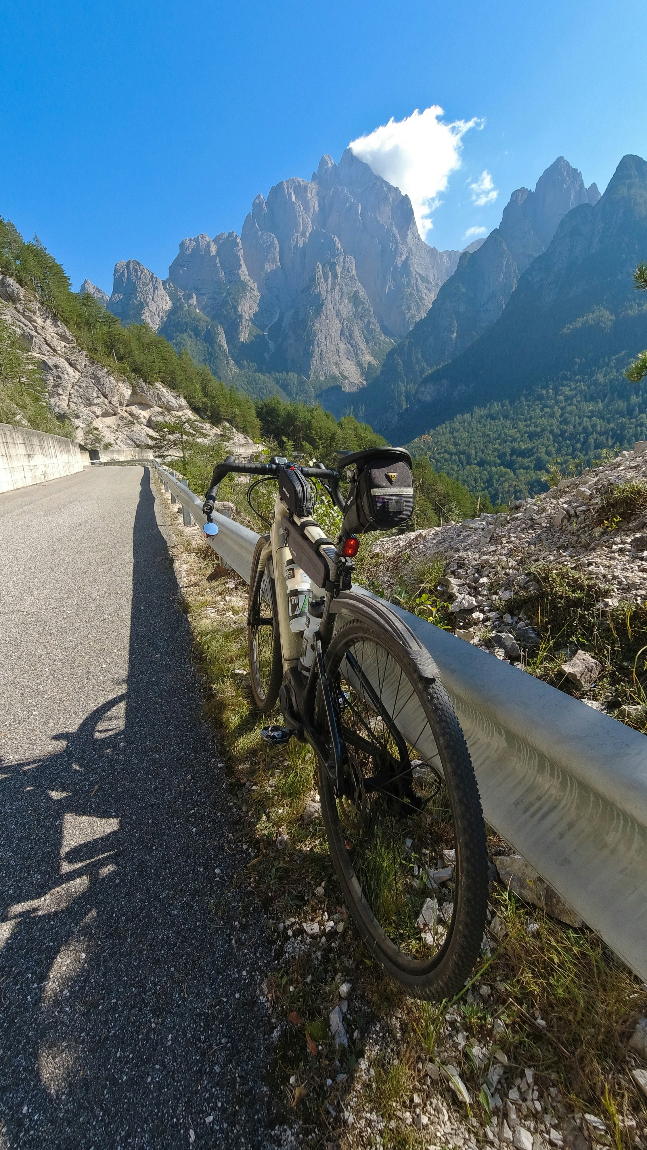 A bicycle rests against a guardrail beside a winding road, framed by towering mountains under a clear blue sky.