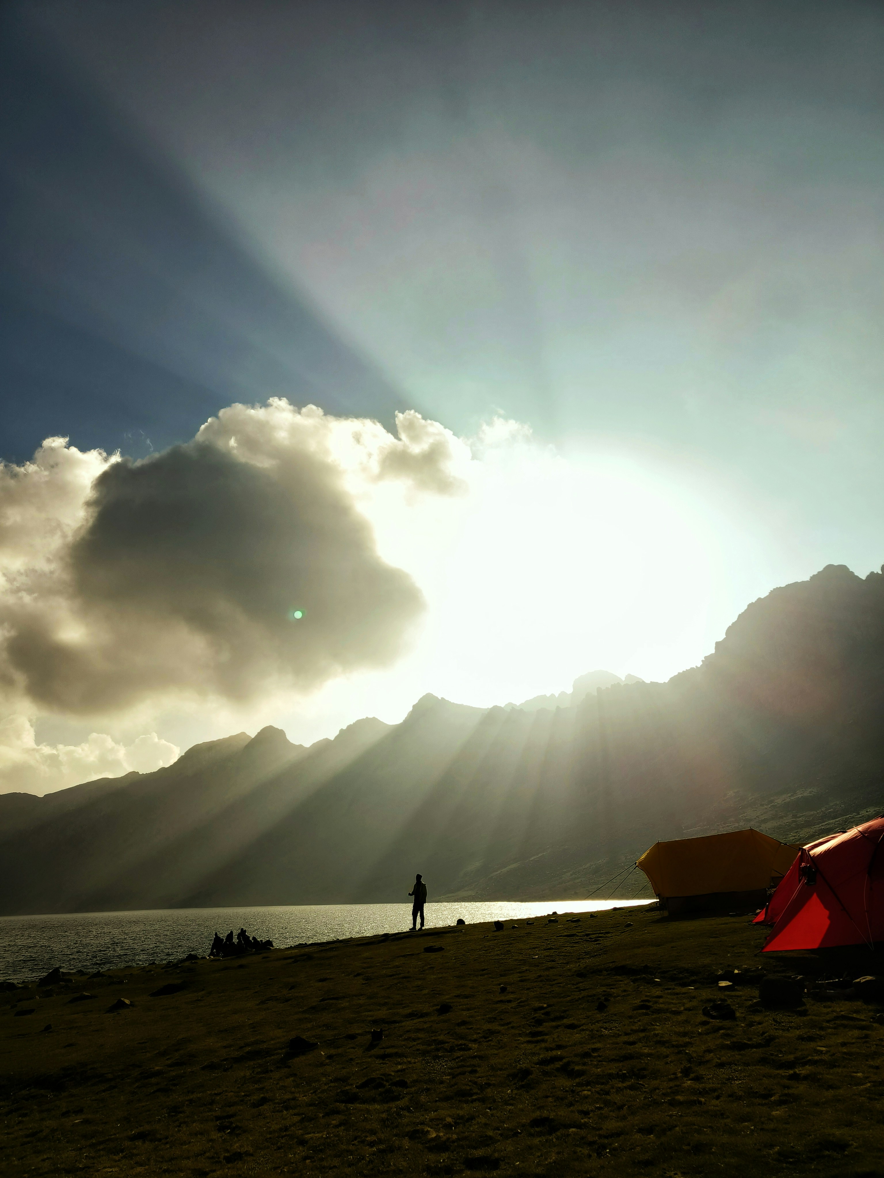 Sun rays slice through the clouds over jagged mountains reflected on a calm lake, with a lone hiker on the shore near red tents.