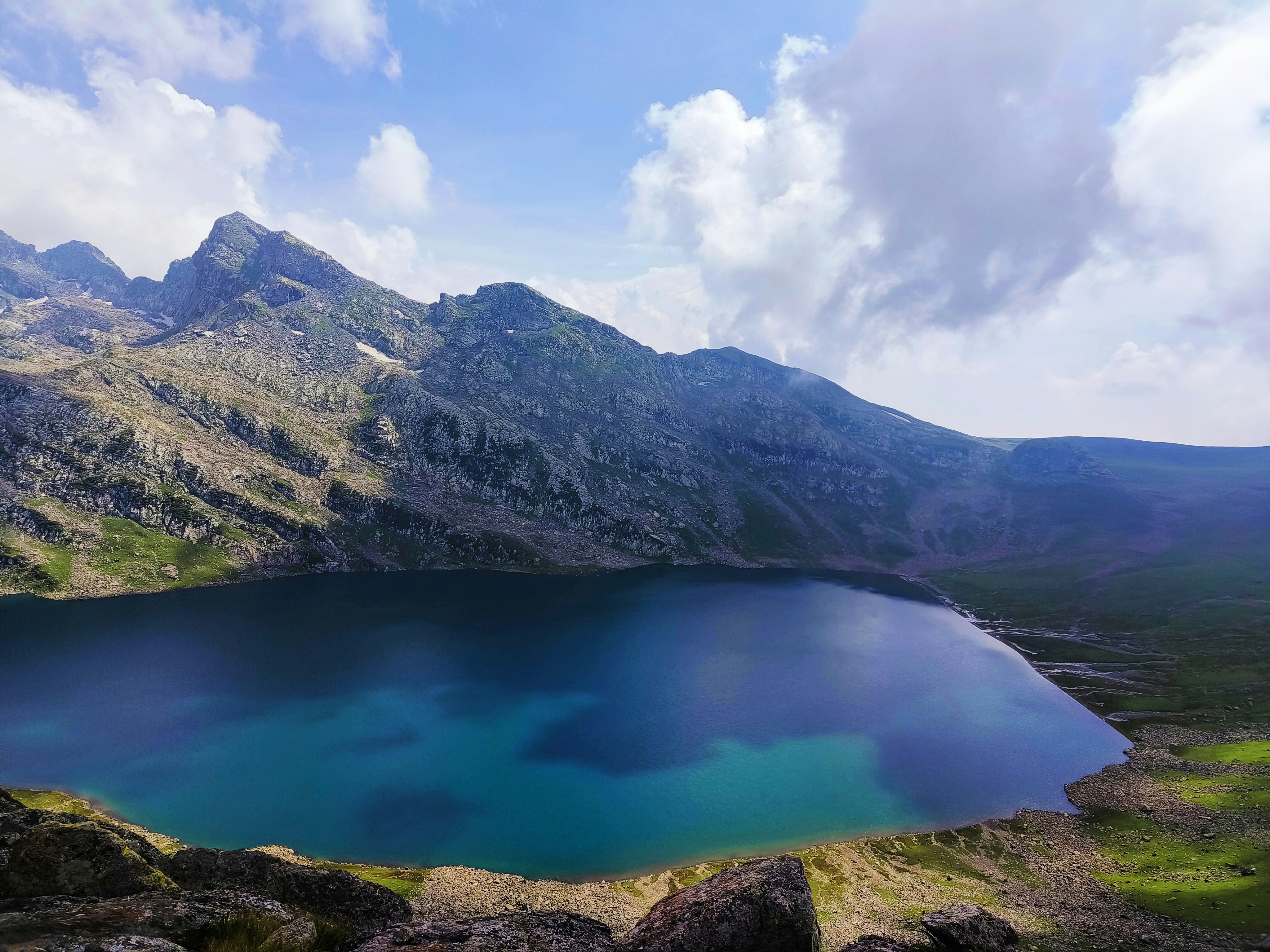 Clear alpine lake with turquoise waters sits in a rugged basin edged by rocky shores and green meadows. Jagged mountains rise around it under a bright, partly cloudy sky.