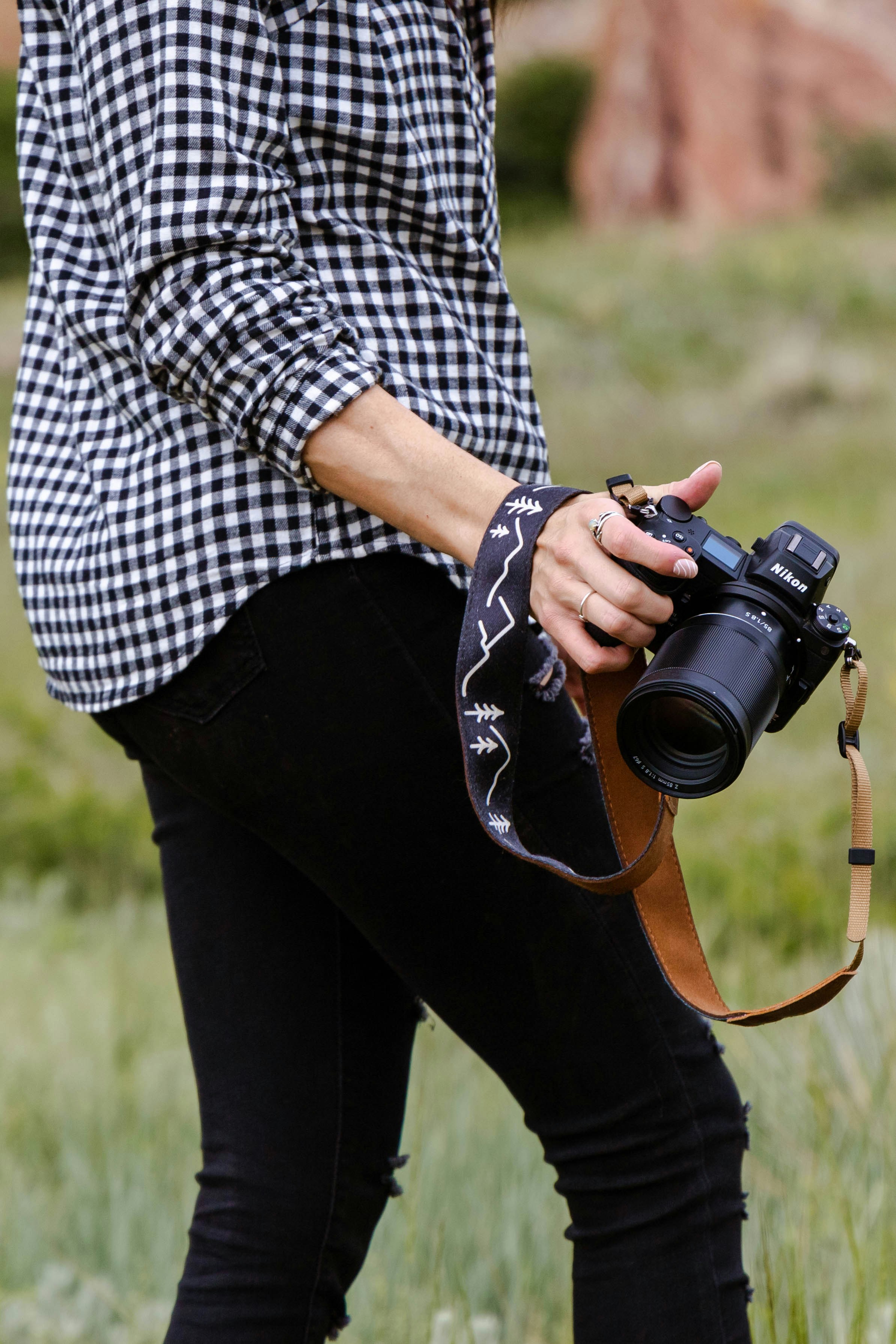 A photographer stands poised in a natural setting, holding a camera with a stylish strap. The focus is on their attire and the equipment, highlighting the connection between the artist and their craft.