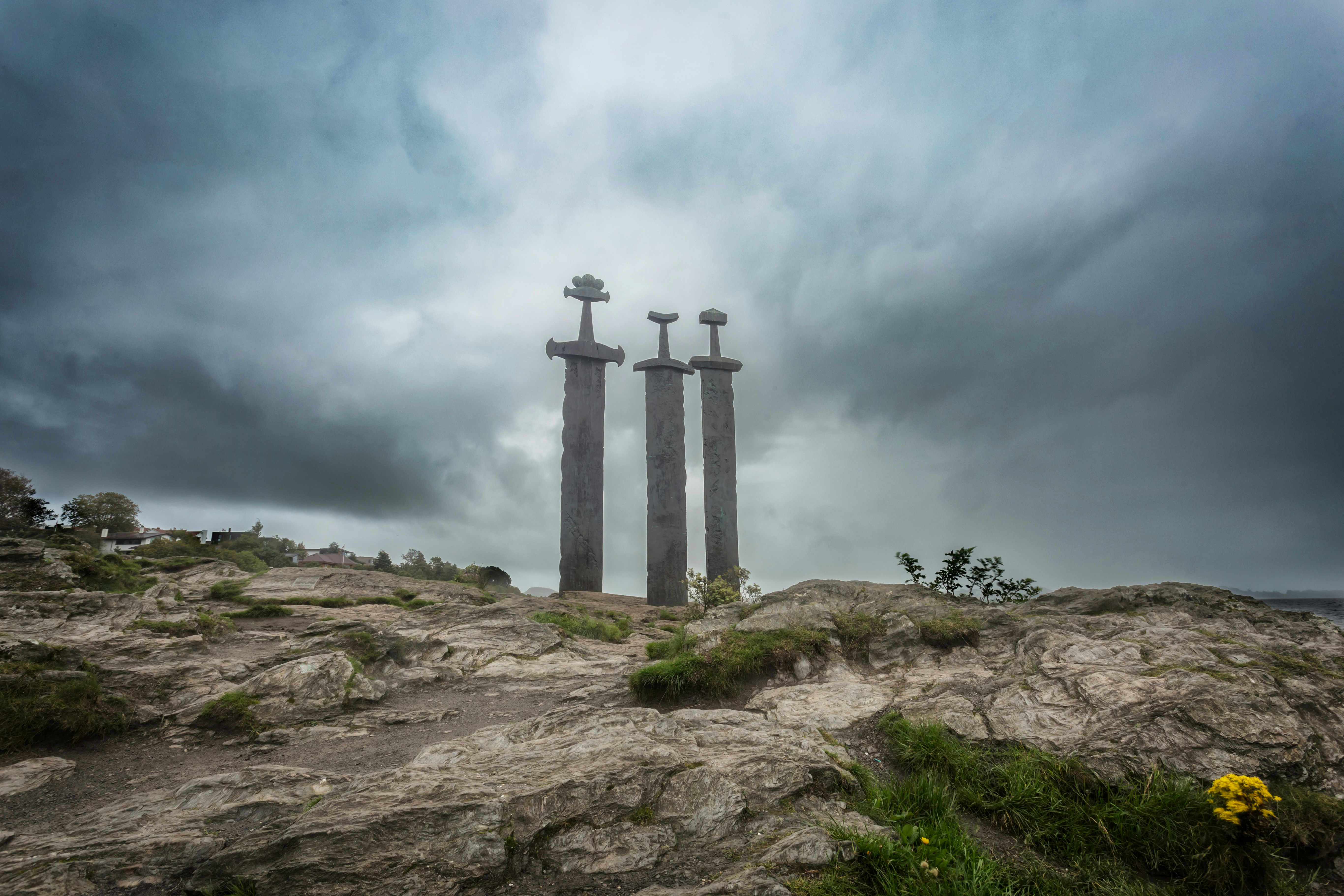 Three towering sword sculptures rise from a rocky hill under a dramatic, cloudy sky.