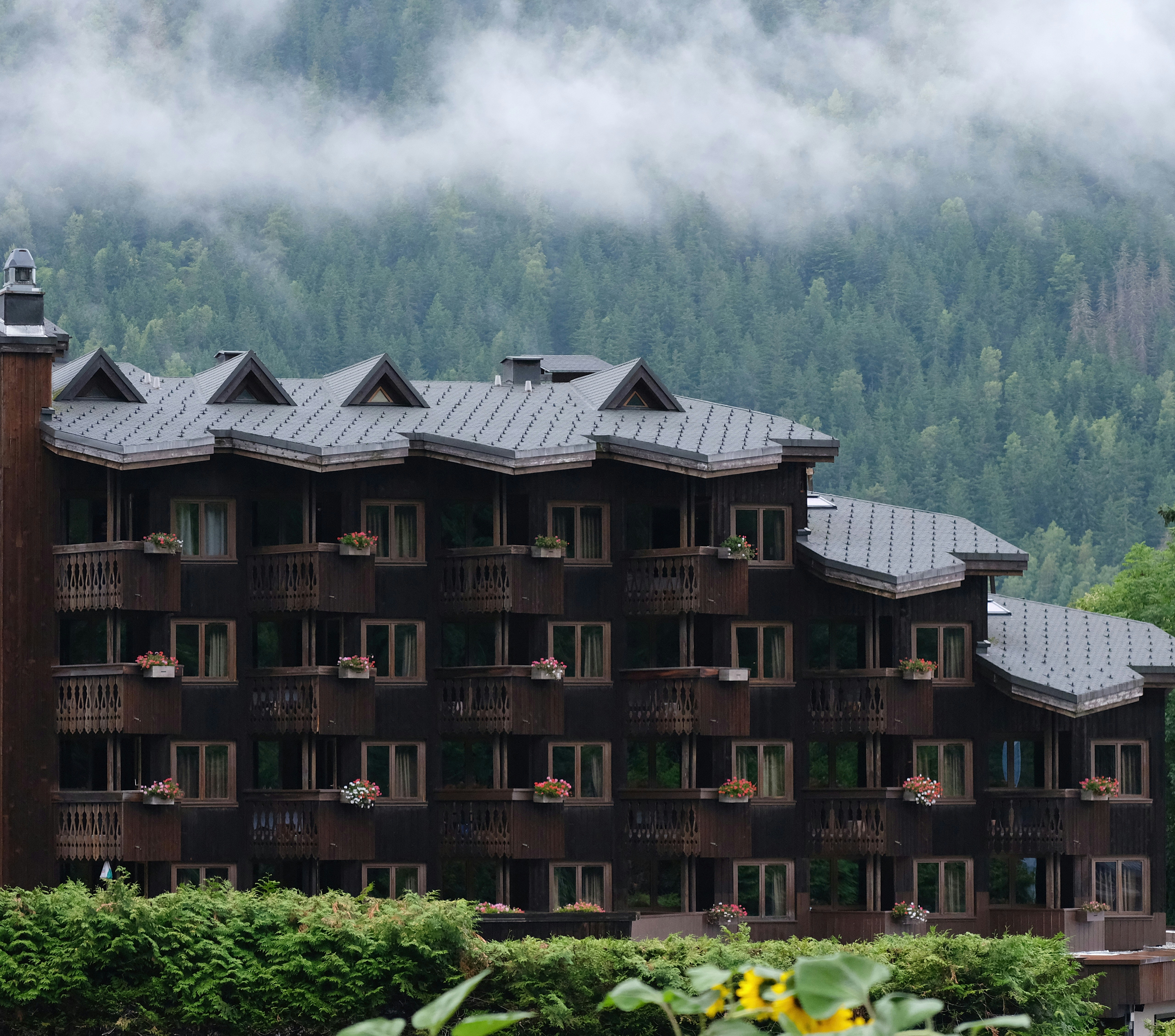 Multi-story dark-wood chalet with staggered gabled roofs and balconies sits along a hillside, backed by misty mountains. Bright sunflowers frame the foreground, adding color.