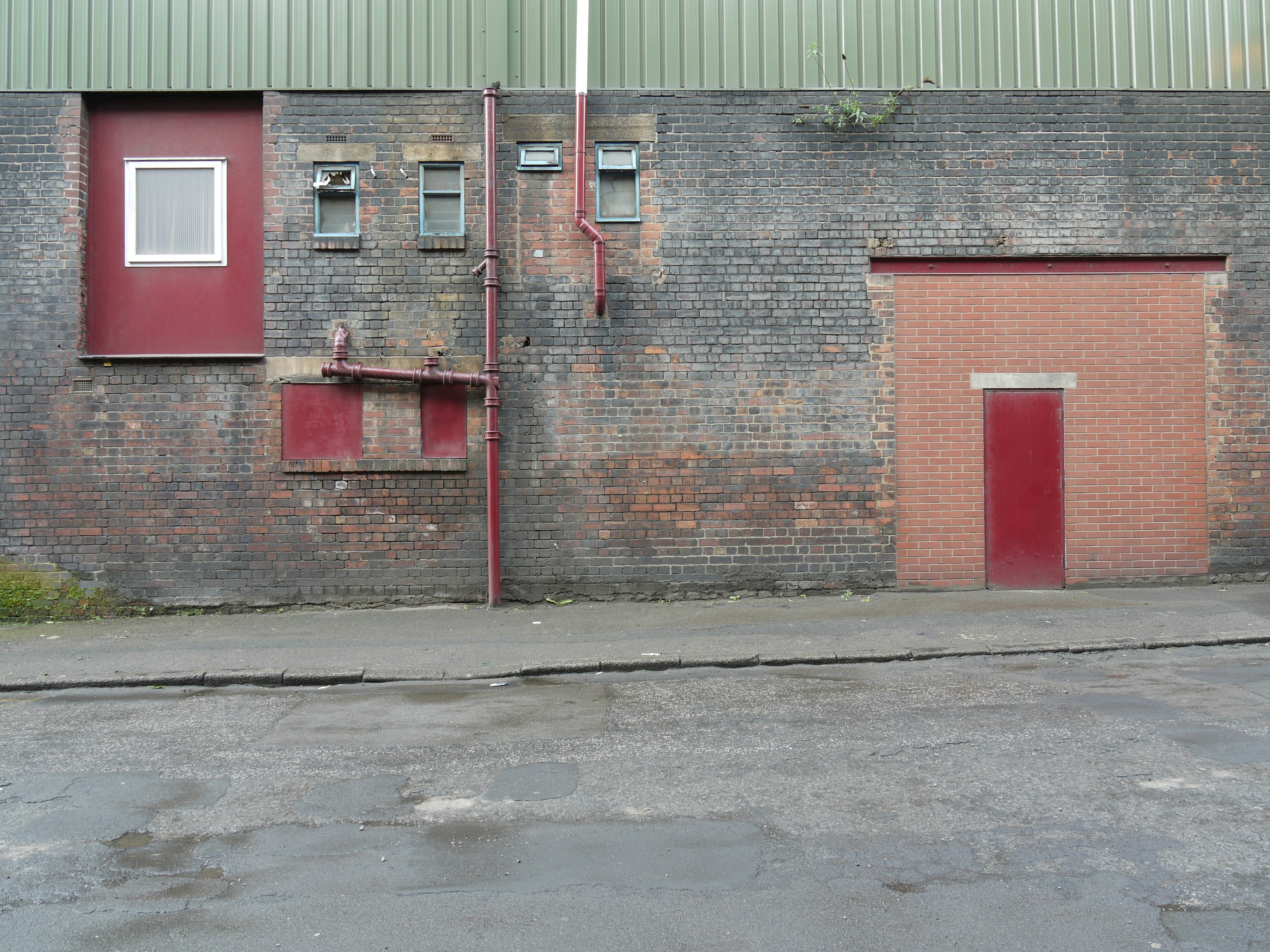 Weathered brick façade with red doors and exposed piping along a quiet street; the scene emphasizes industrial textures and muted color contrasts.