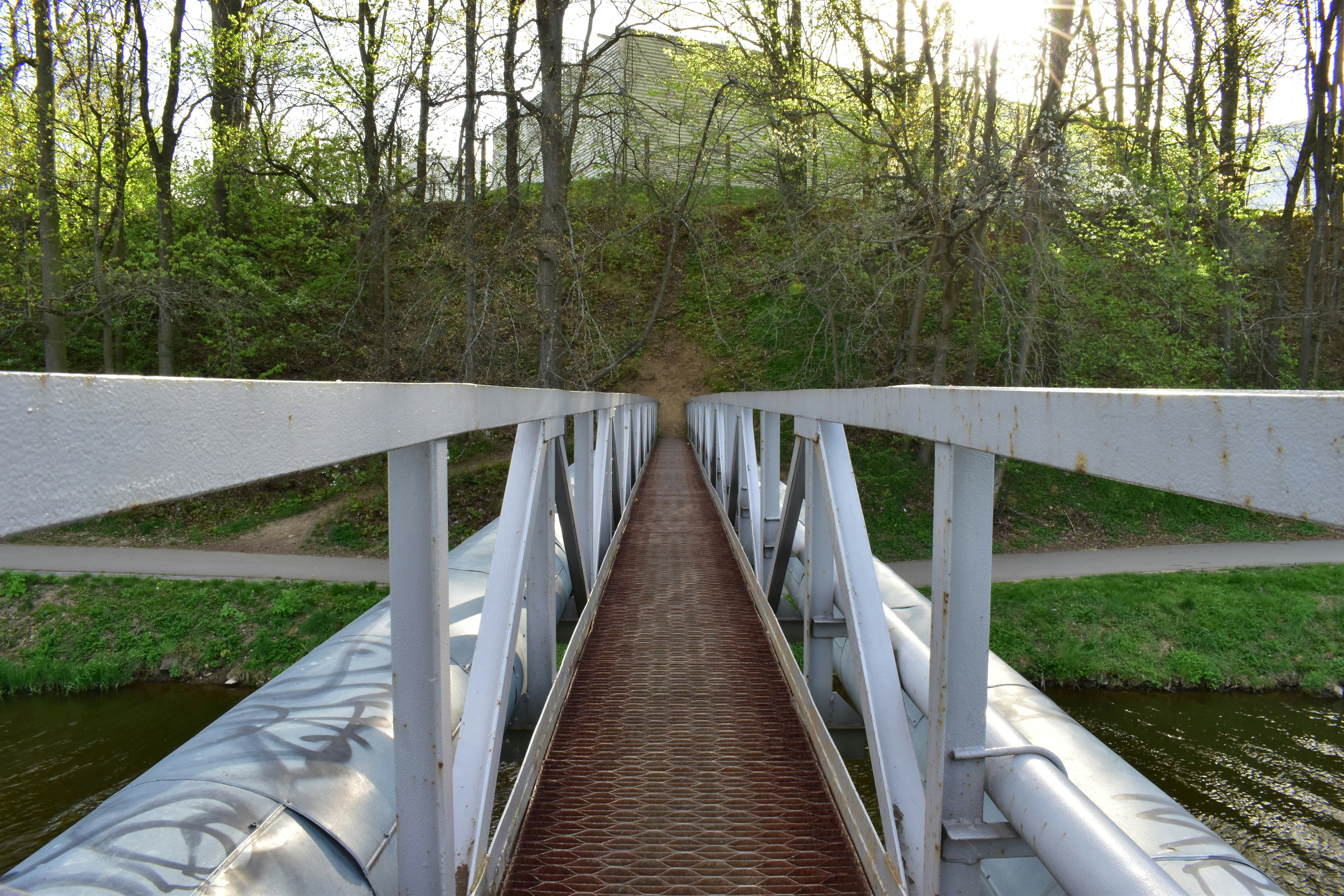 Metal bridge spanning a serene waterway, leading towards a lush green hillside under soft sunlight.