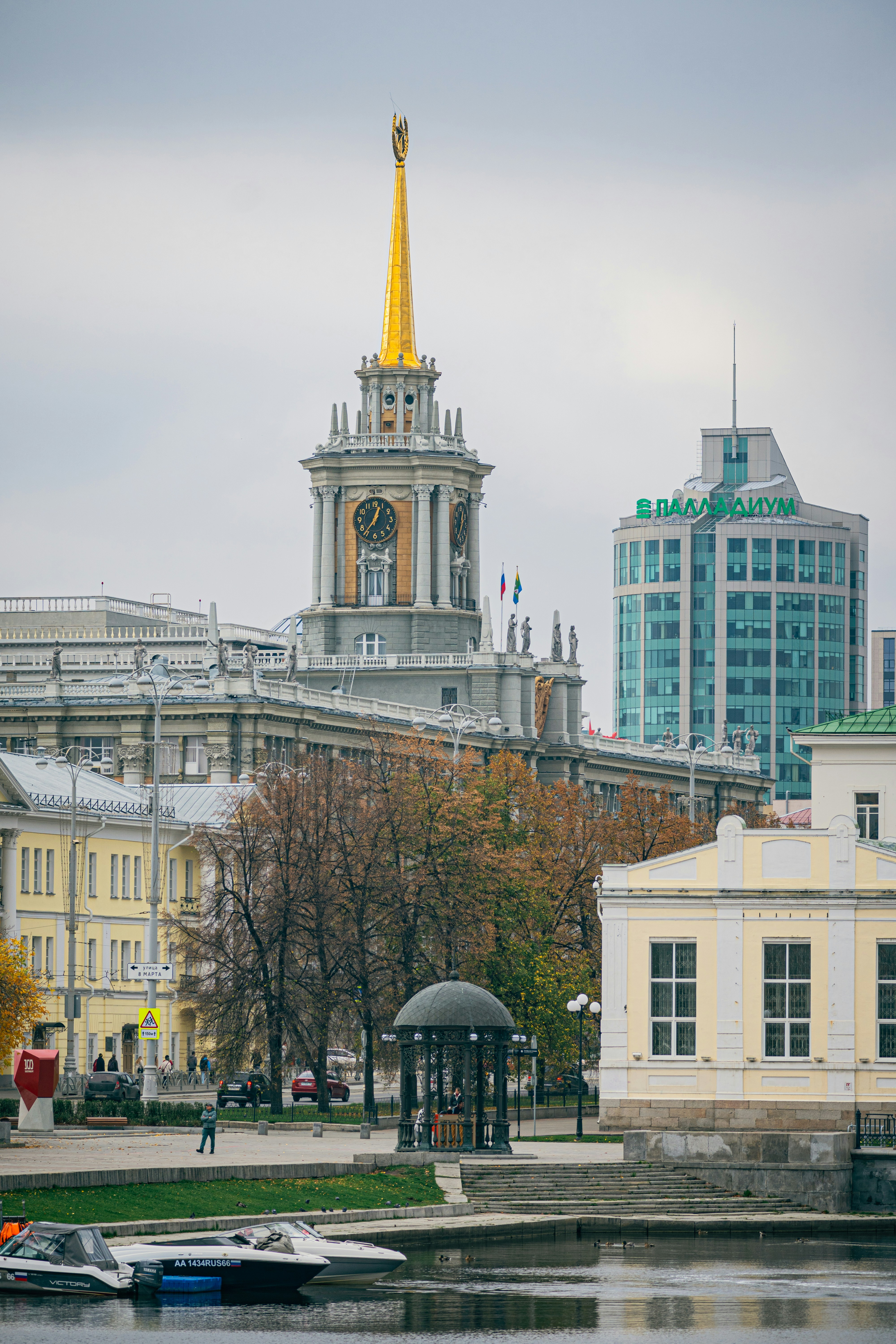 Golden spire atop a historical building overlooking a serene river, framed by modern architecture and autumn foliage.