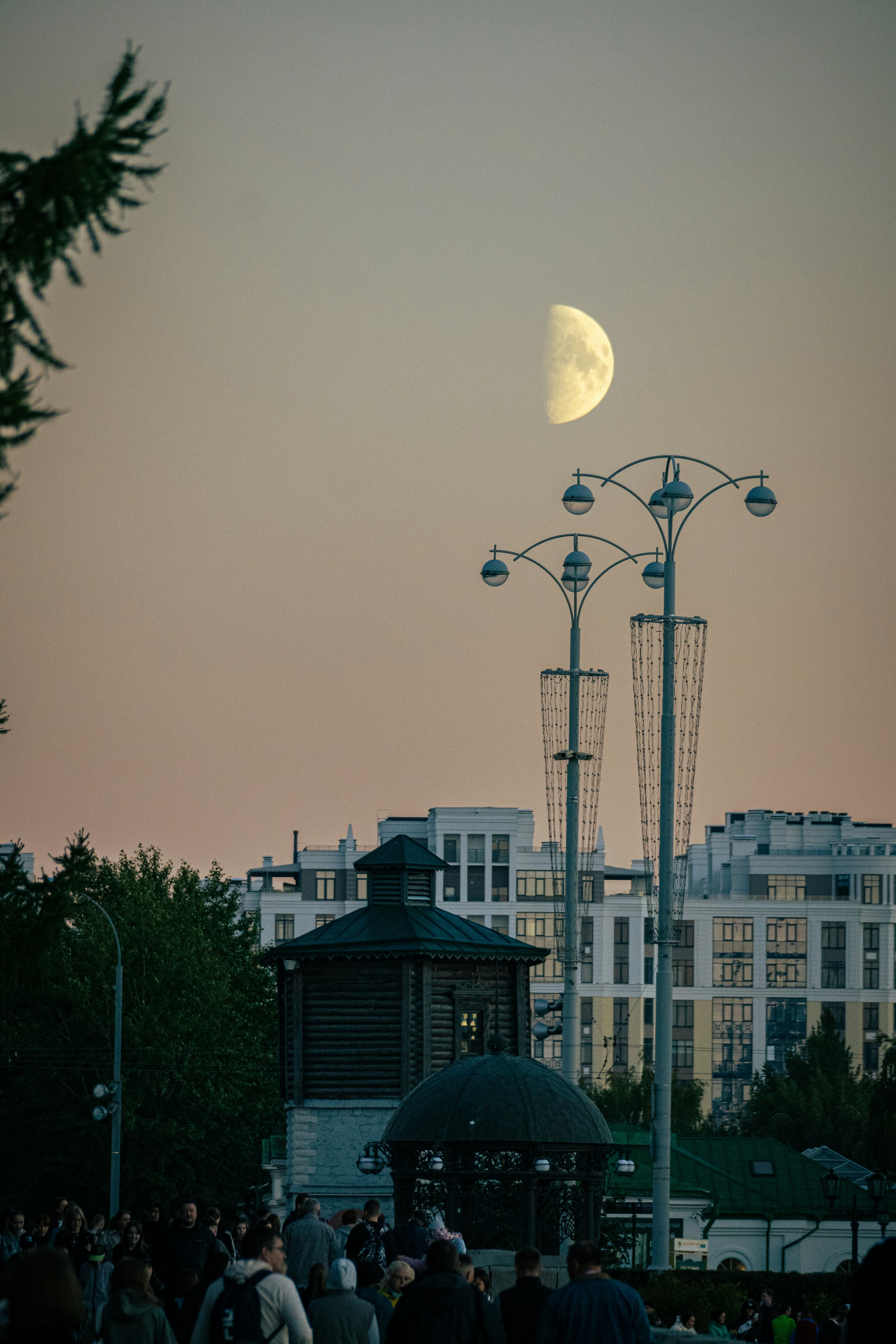 Moonrise Over a City Park: Dusk Light, Glass-Lamp Arches, and a Crowd