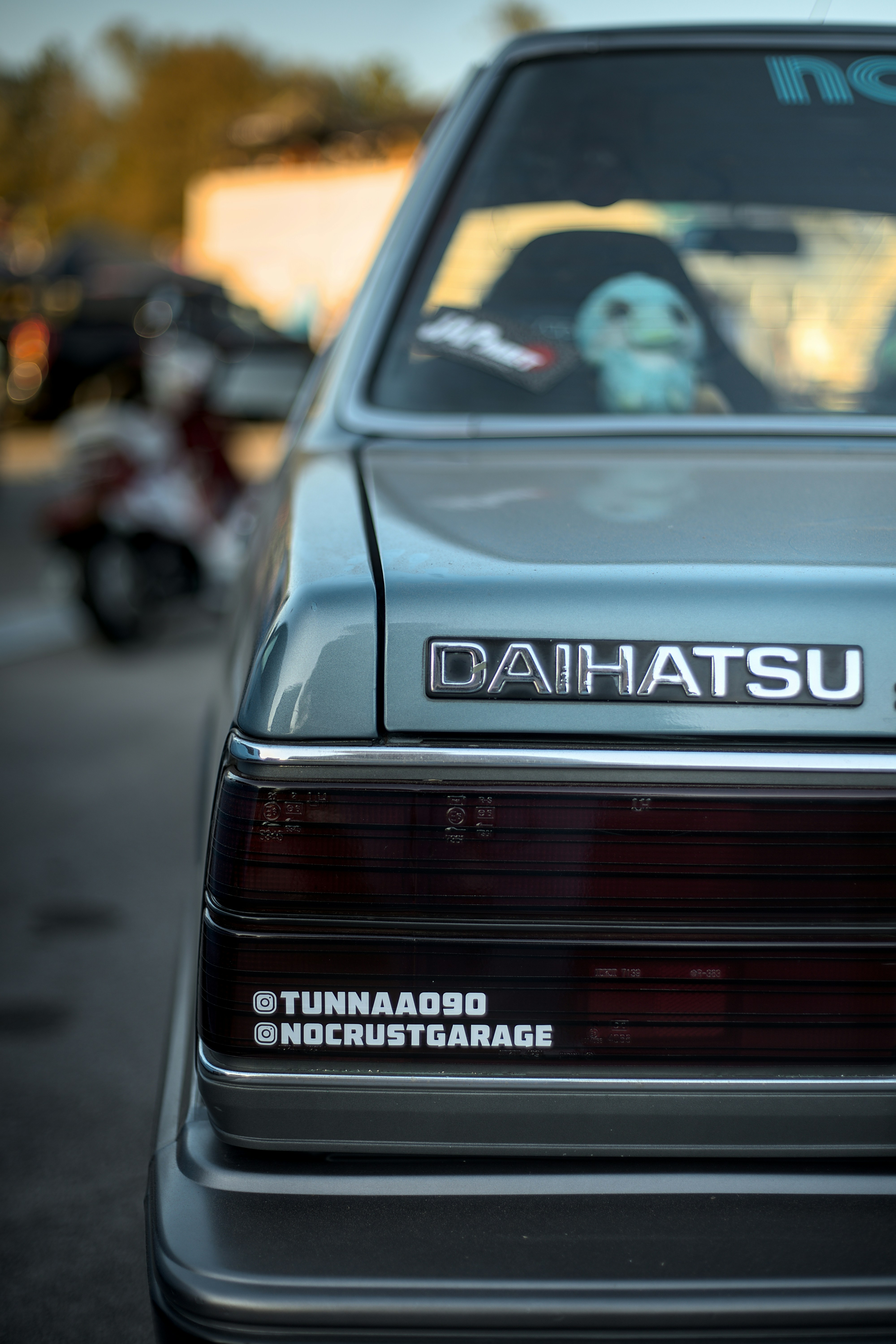 Tailgate Portrait: Daihatsu Emblem Amid Stickered Trunk and Plush in Sunset Blur
