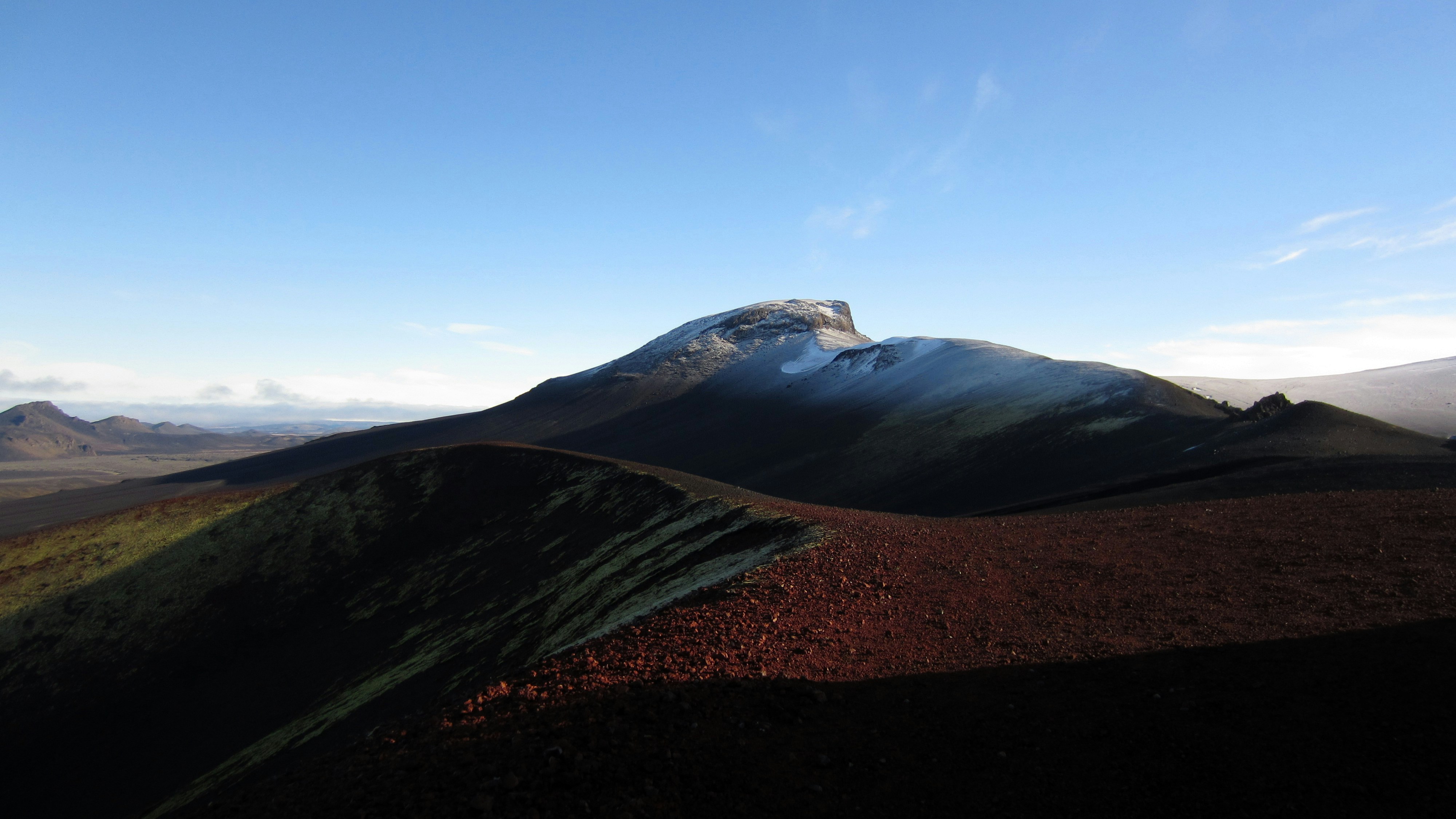 Volcanic ridge rises toward a distant summit under a clear blue sky. Foreground shows dark red-brown soil and mossy slopes.
