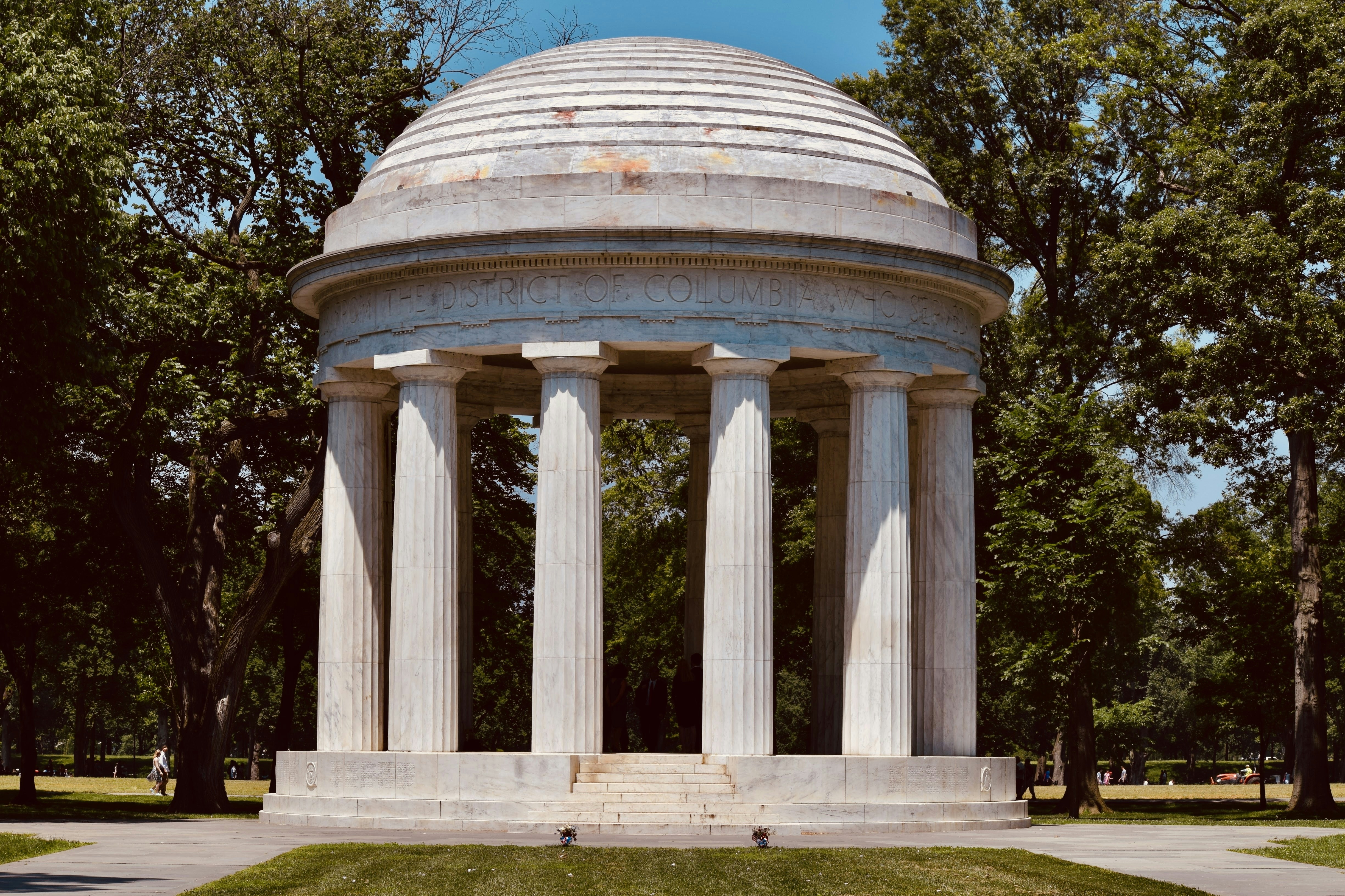 A classical pavilion with a domed roof and fluted columns, surrounded by lush greenery in a public park.