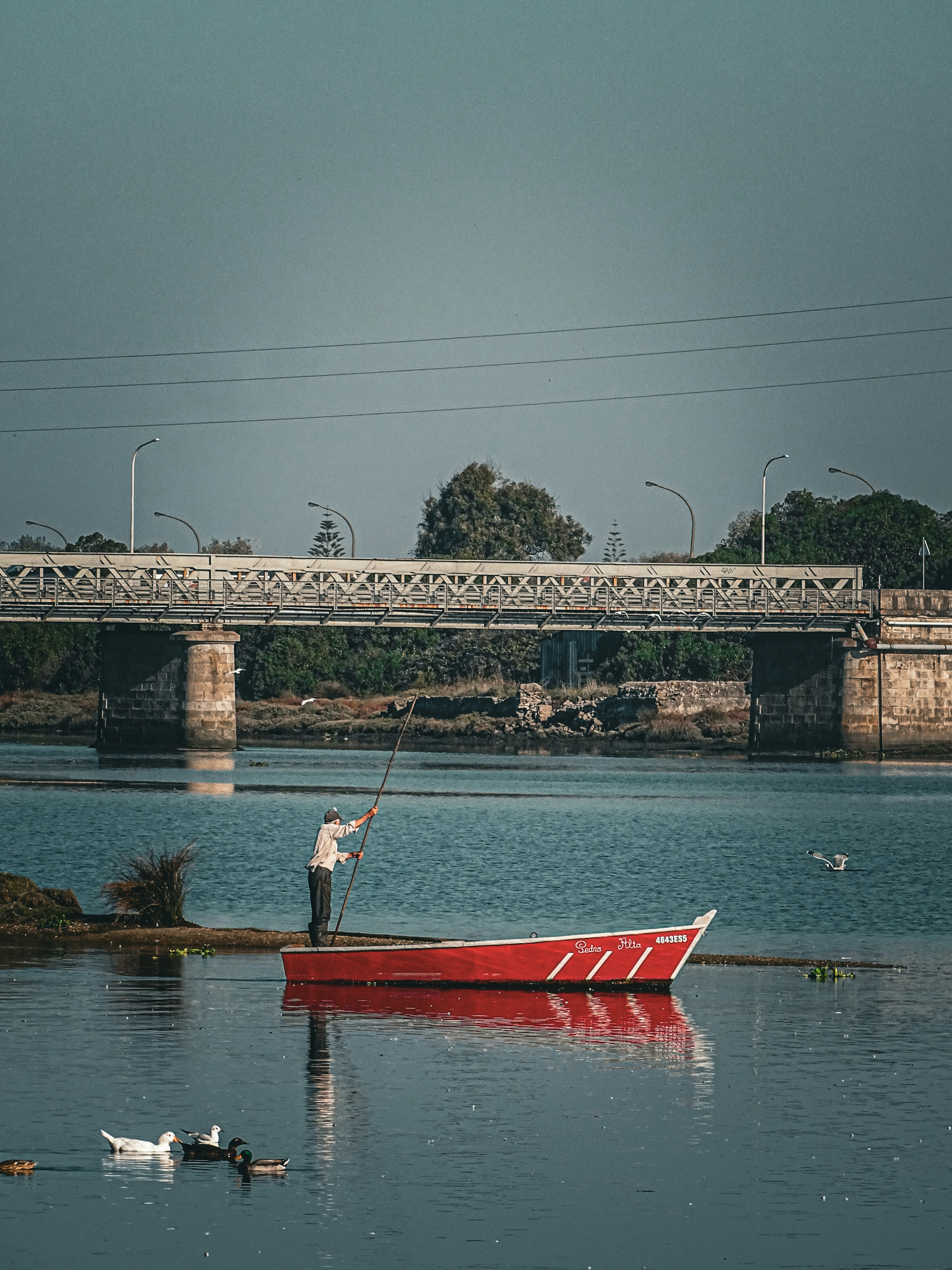 A person stands on a red boat fishing along a calm river with a steel truss bridge spanning the background.