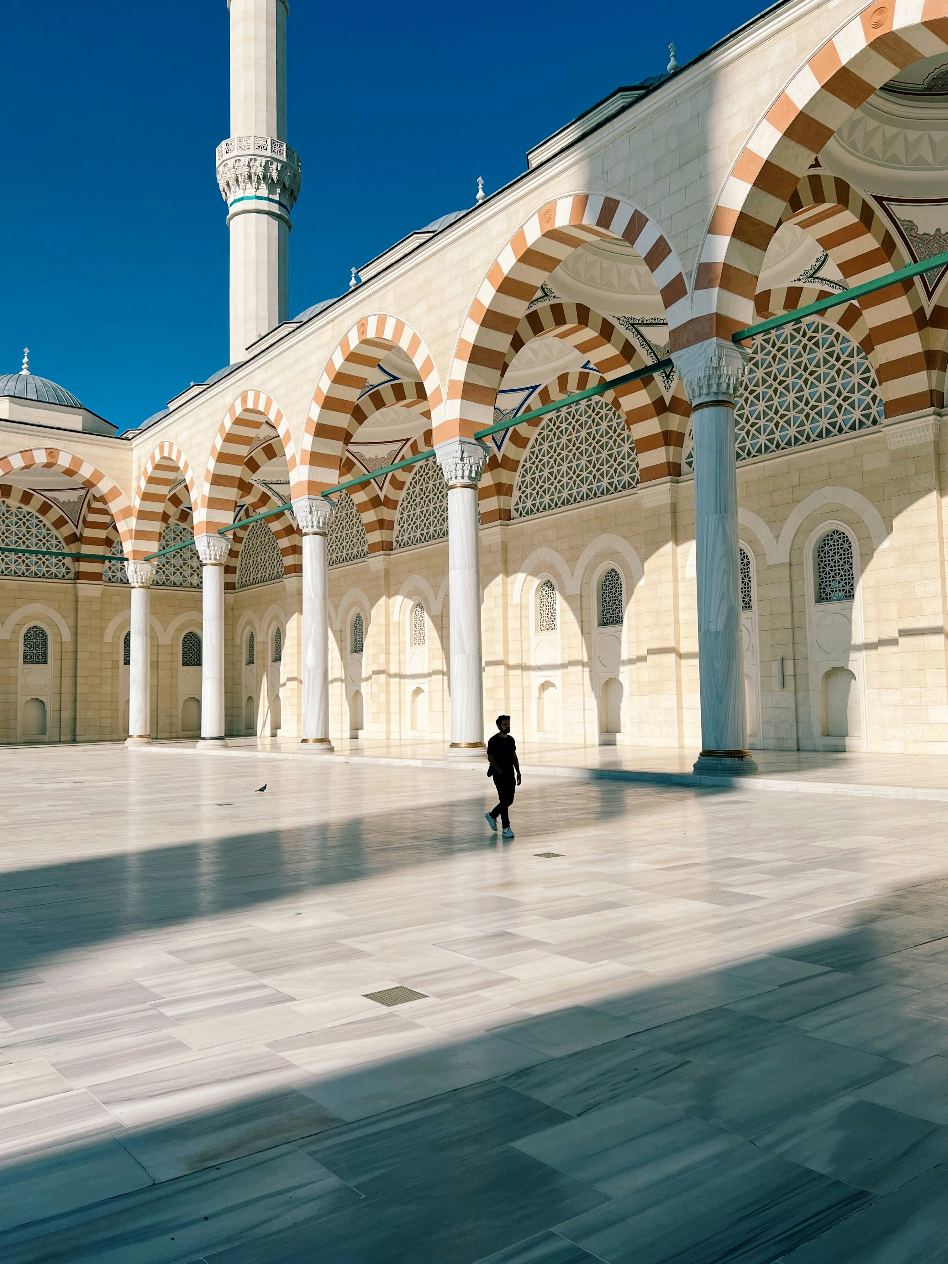 Person walks through the sunlit courtyard of Çamlıca Mosque, framed by grand arches and a clear blue sky.