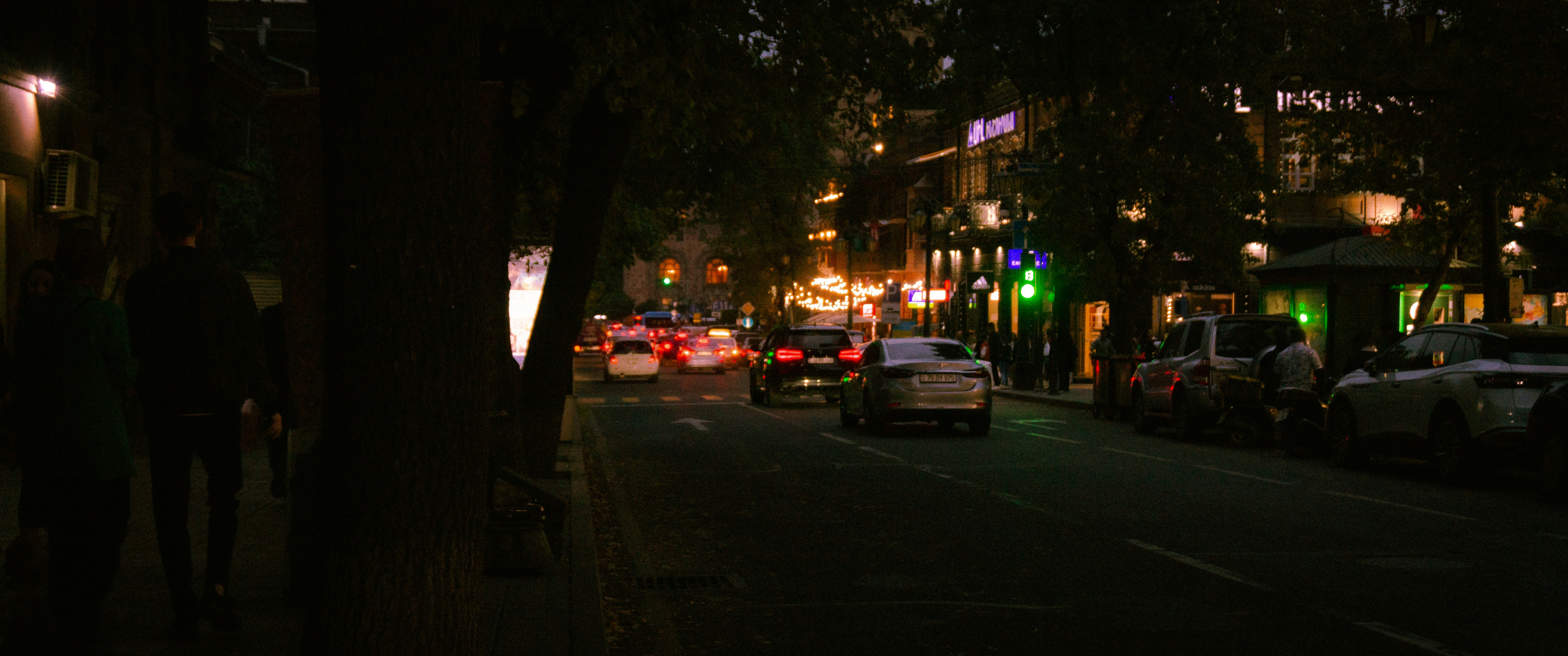 Dimly lit urban street with parked cars and warm glowing lights from nearby buildings.