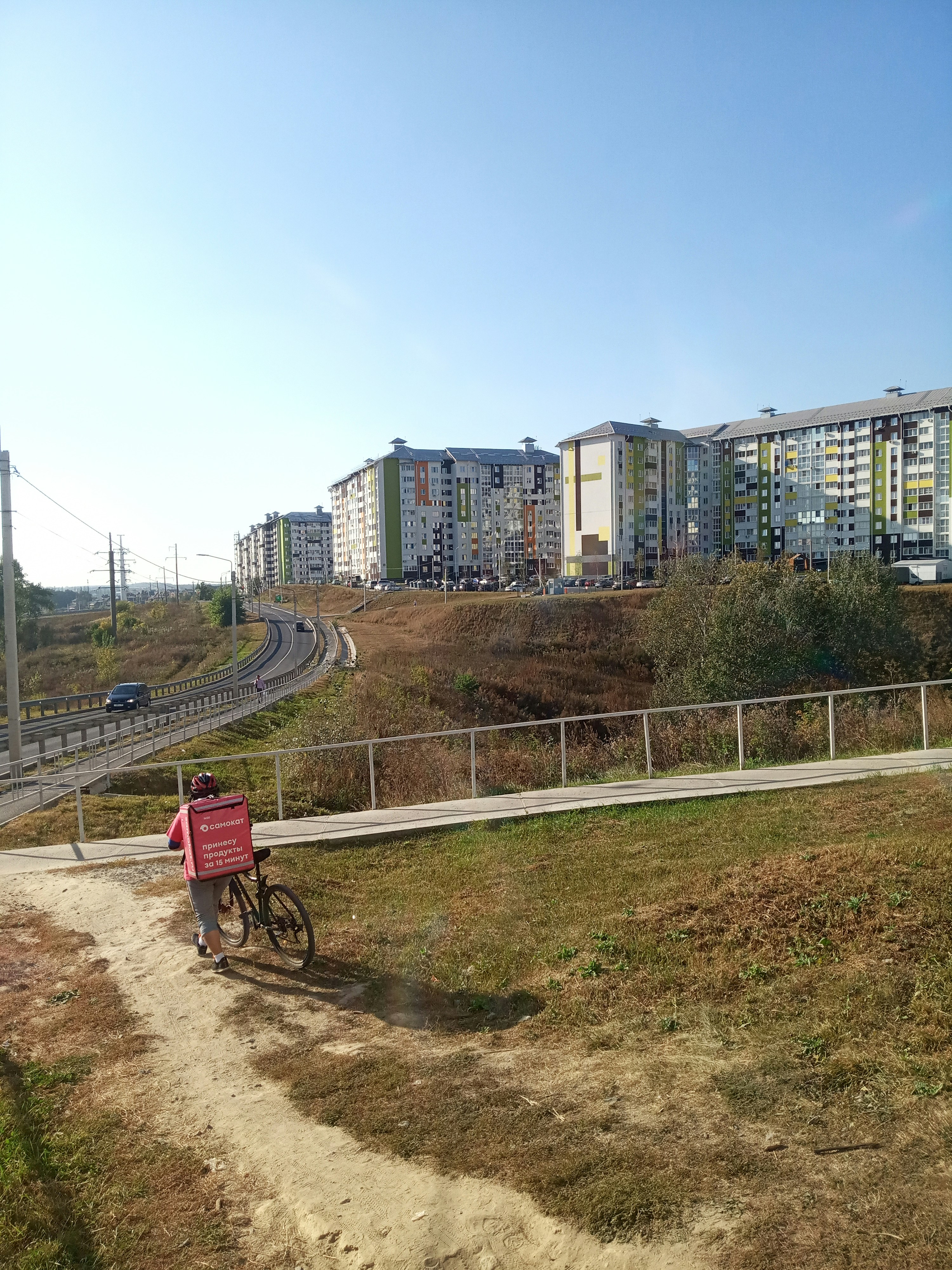 Photograph of a cyclist with a bike on a dirt path beside a railing, with a curving road and a row of apartment buildings on a hillside in the background.