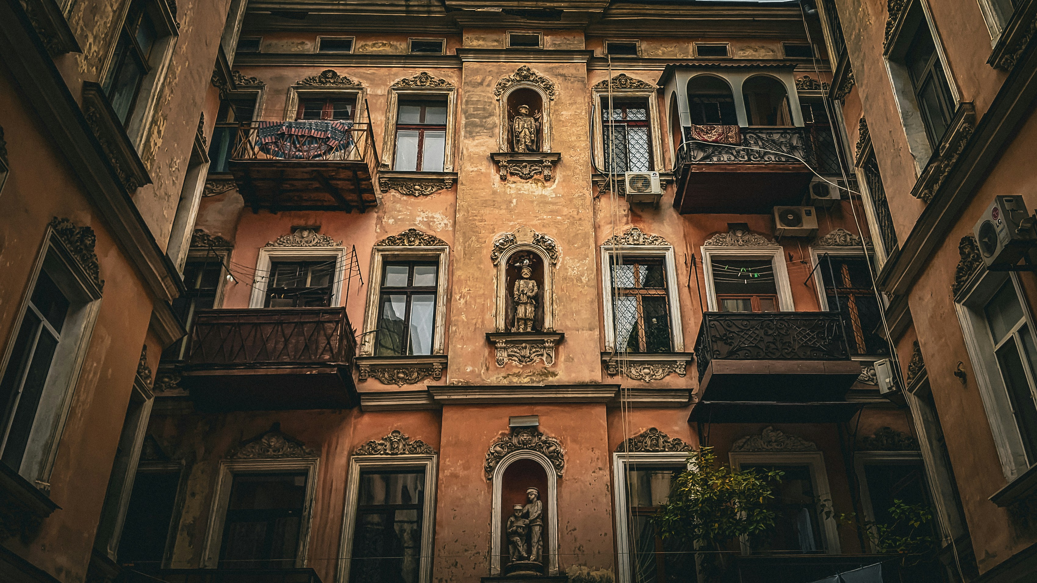 Weathered European courtyard facade with warm orange plaster and multiple balconies; a central niche statue anchors the symmetry of the masonry.