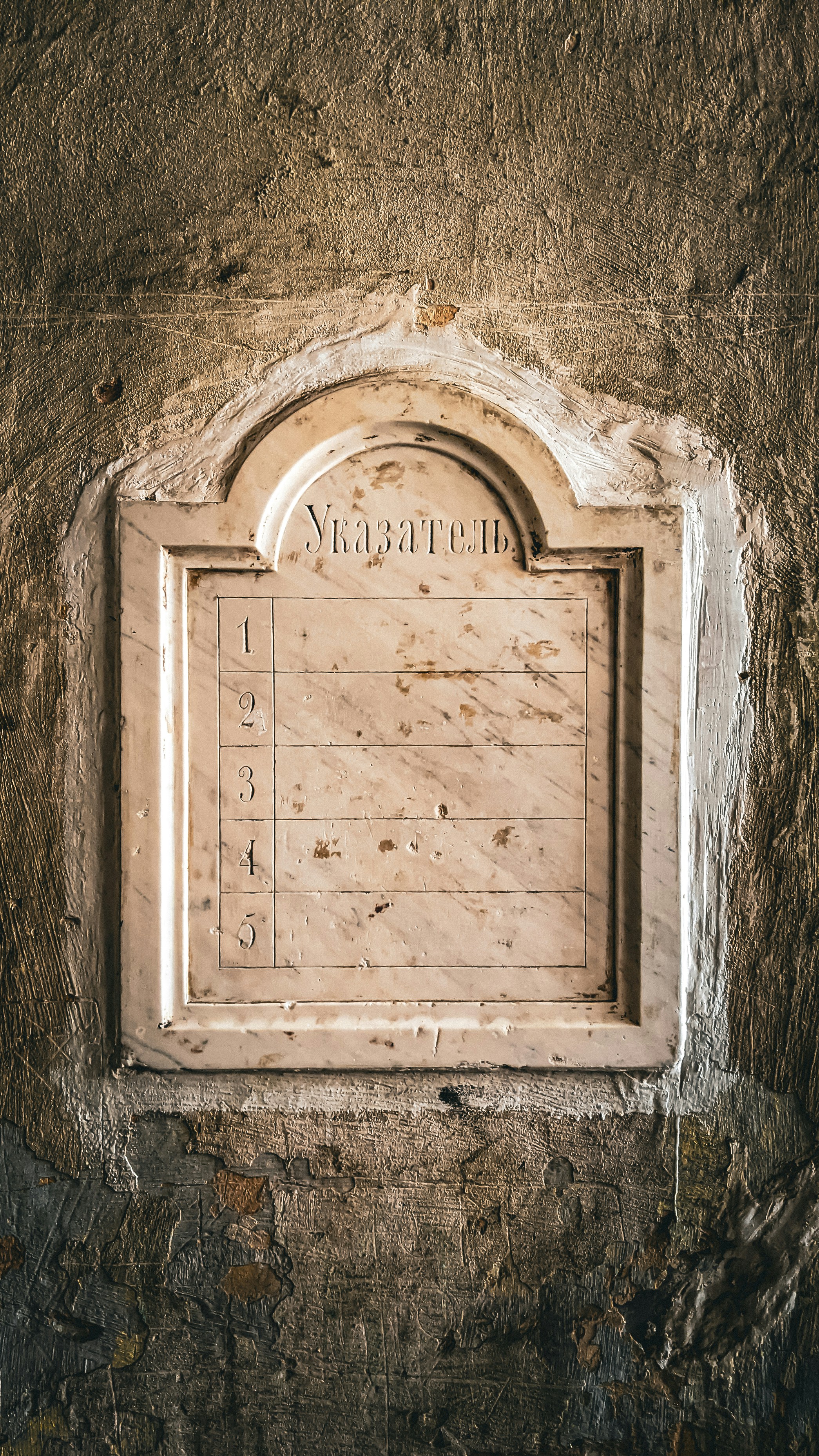 A weathered plaster-framed signboard embedded in a rough, peeling wall; faded lines on the panel hint at a once-clear schedule.
