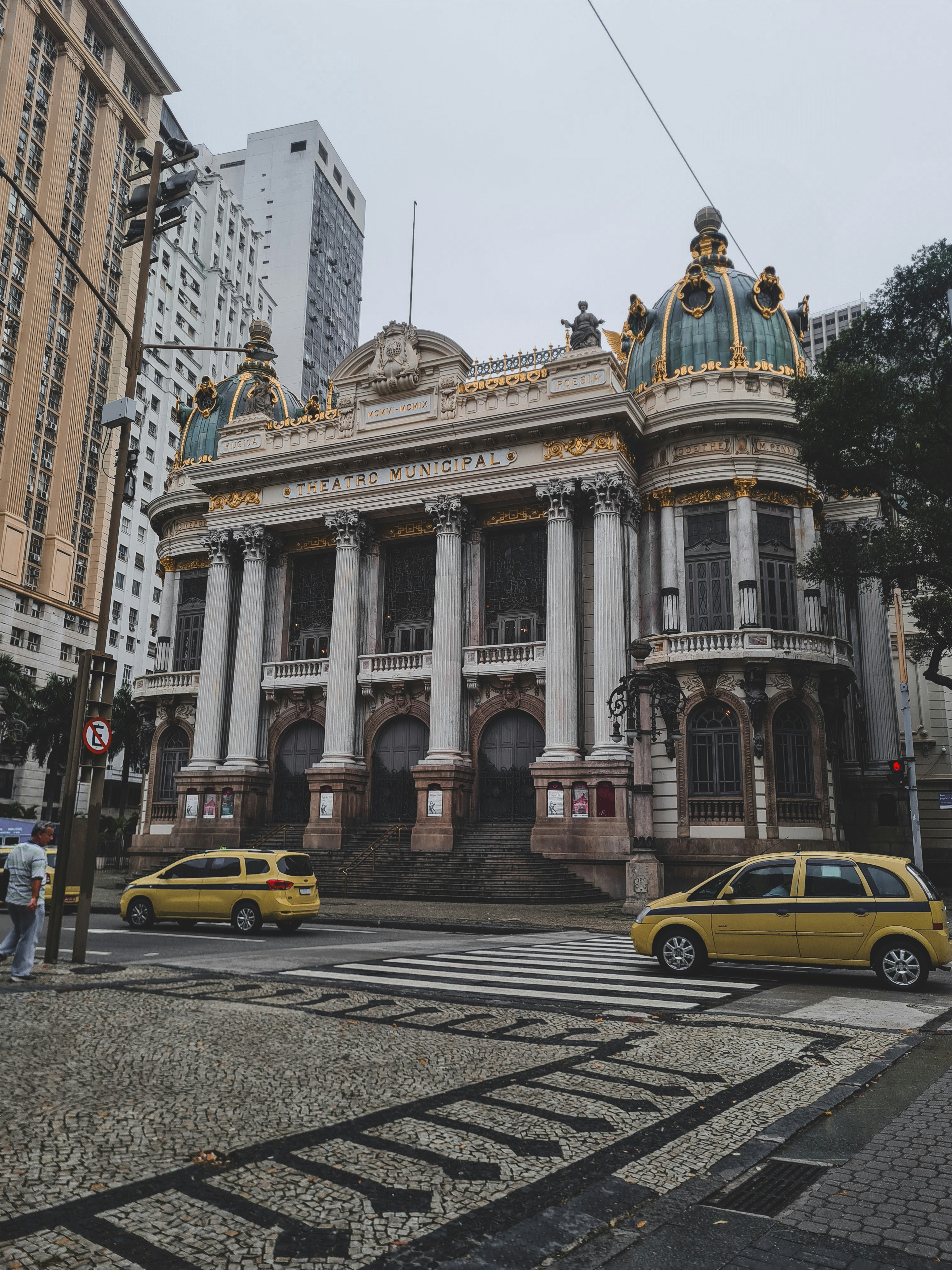 Ornate historic building with columns and domed towers, flanked by modern skyscrapers and yellow taxis on a city street.