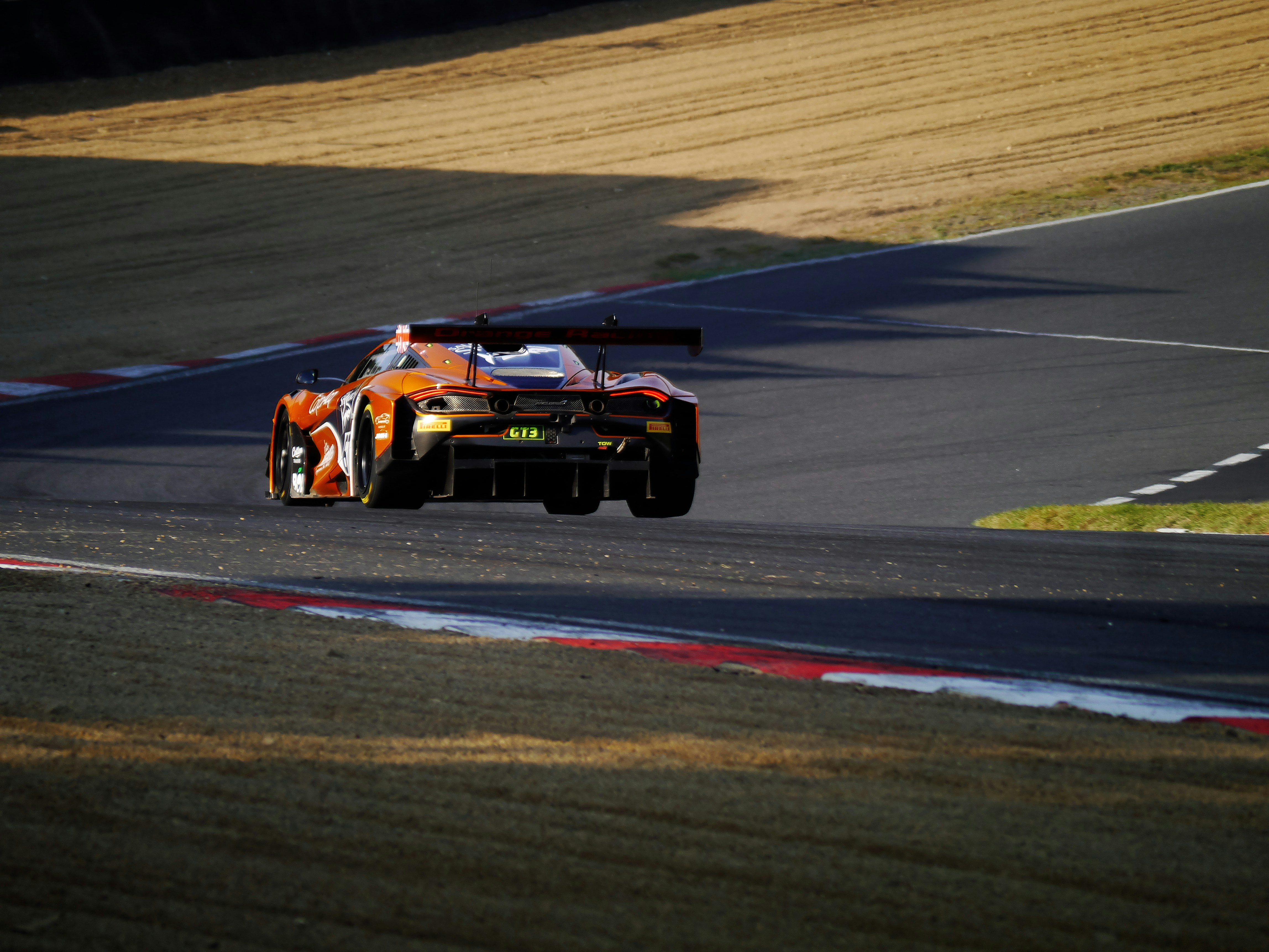 Photograph of an orange race car navigating a sunlit chicane on a race circuit. The shot emphasizes speed, precision, and the car's low, aggressive stance.