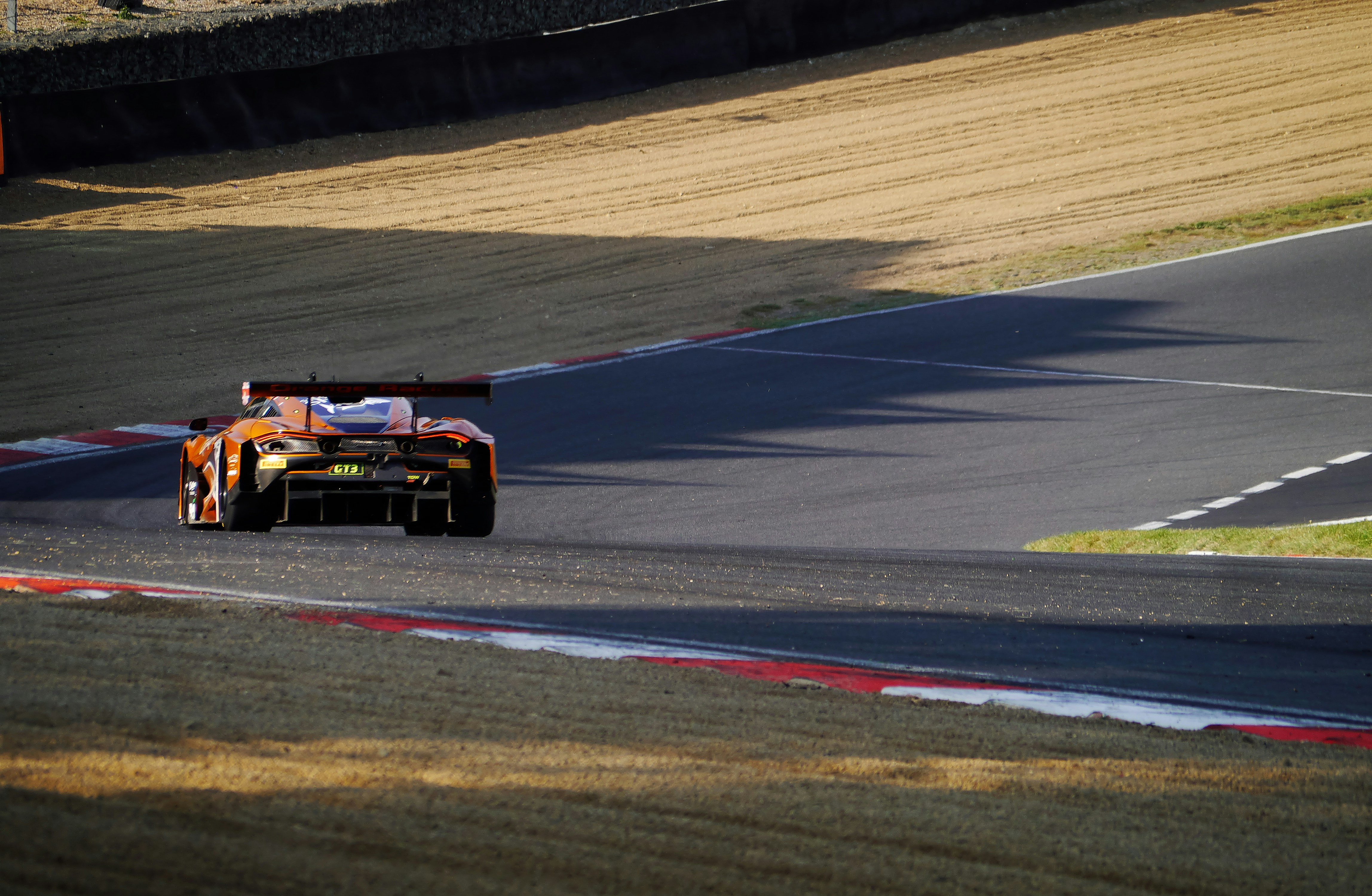 Orange race car tackles a sweeping curve on a sunlit race track.