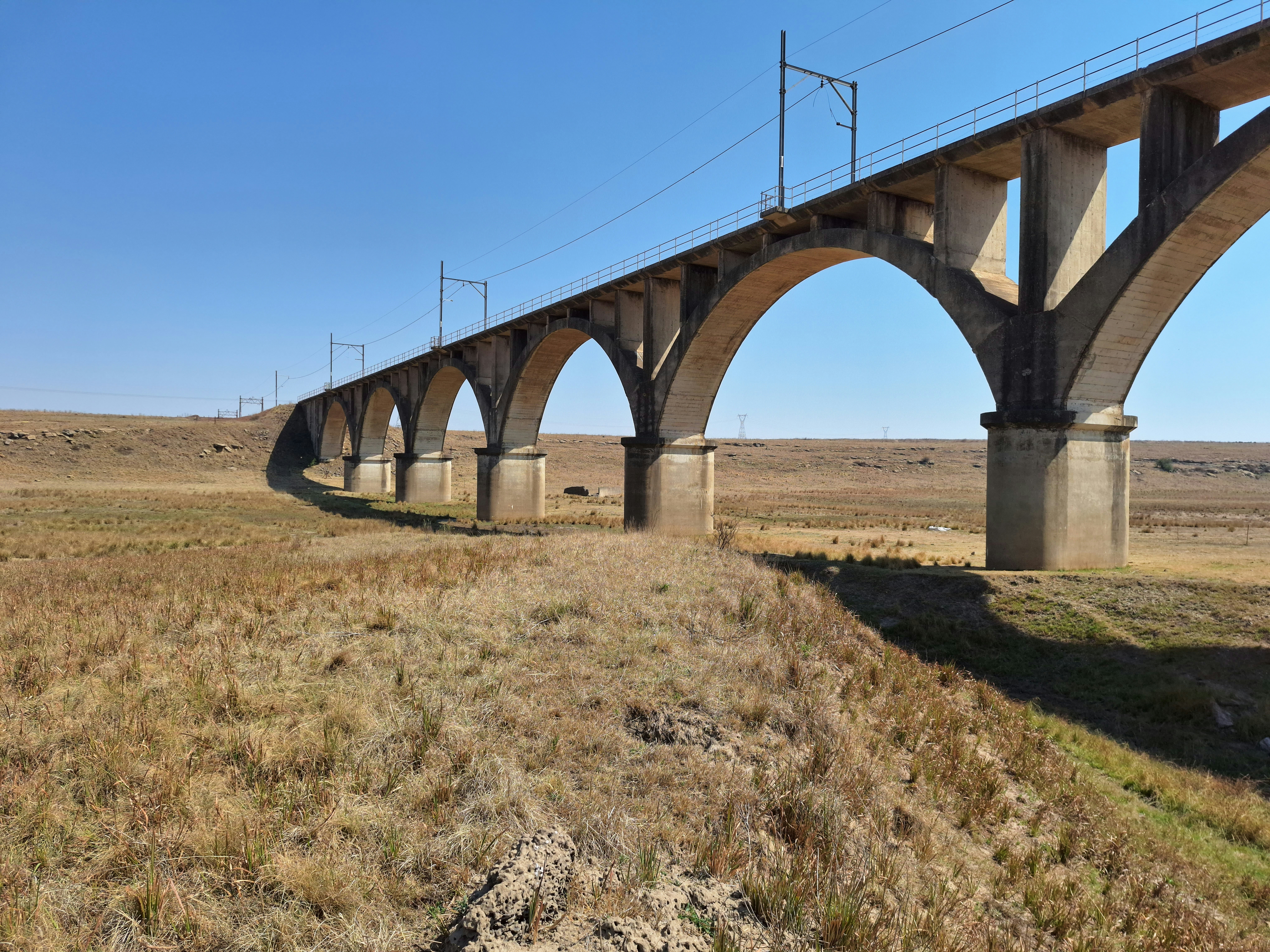Concrete railway arches stretch across a dry, grassy plain under a clear blue sky. The perspective highlights depth as the receding arches guide the eye along the span.