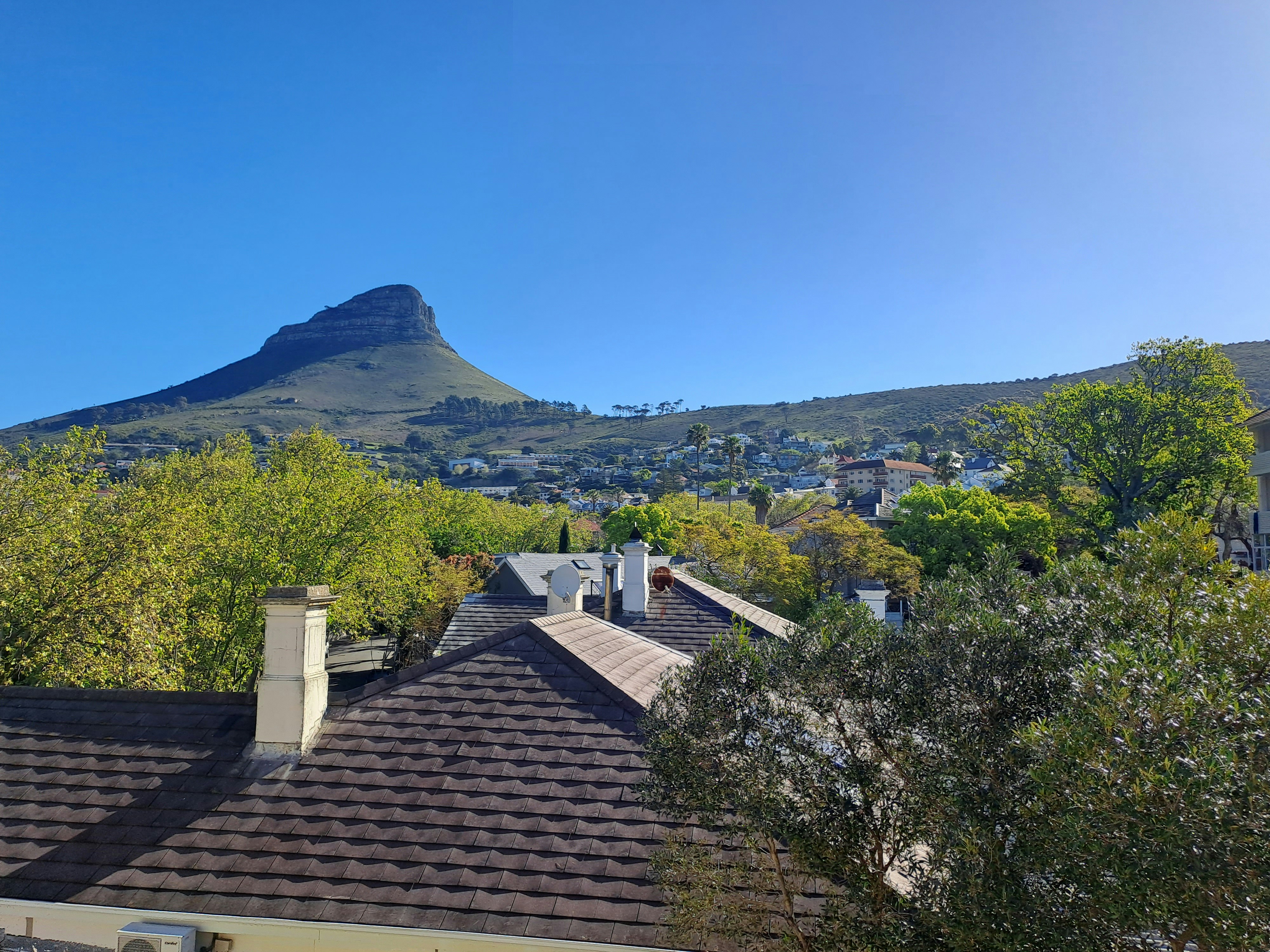 Sunlit mountain peak rises behind a suburban roofscape with tiled roofs and green trees under a clear blue sky.