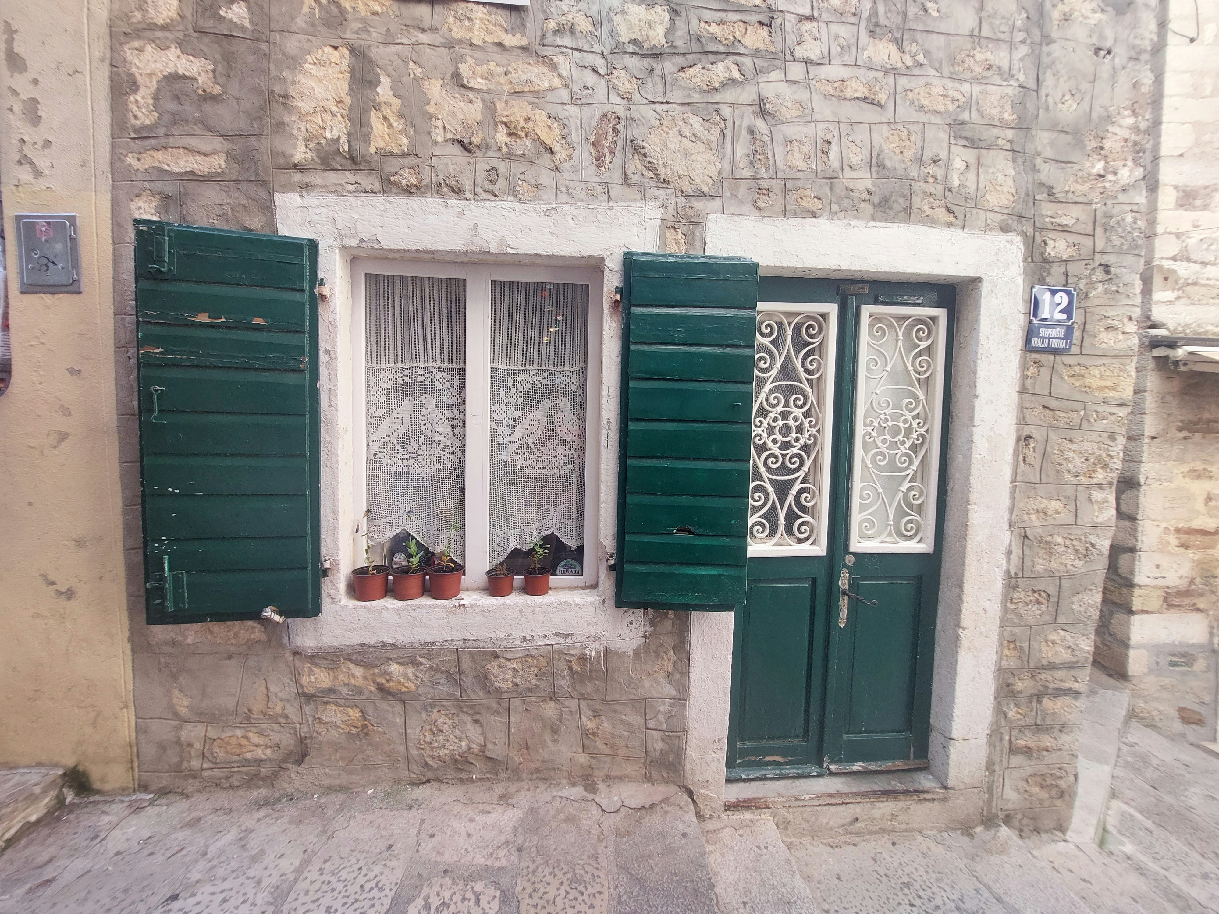 Charming stone facade with green shutters and a decorative door, adorned with potted plants.