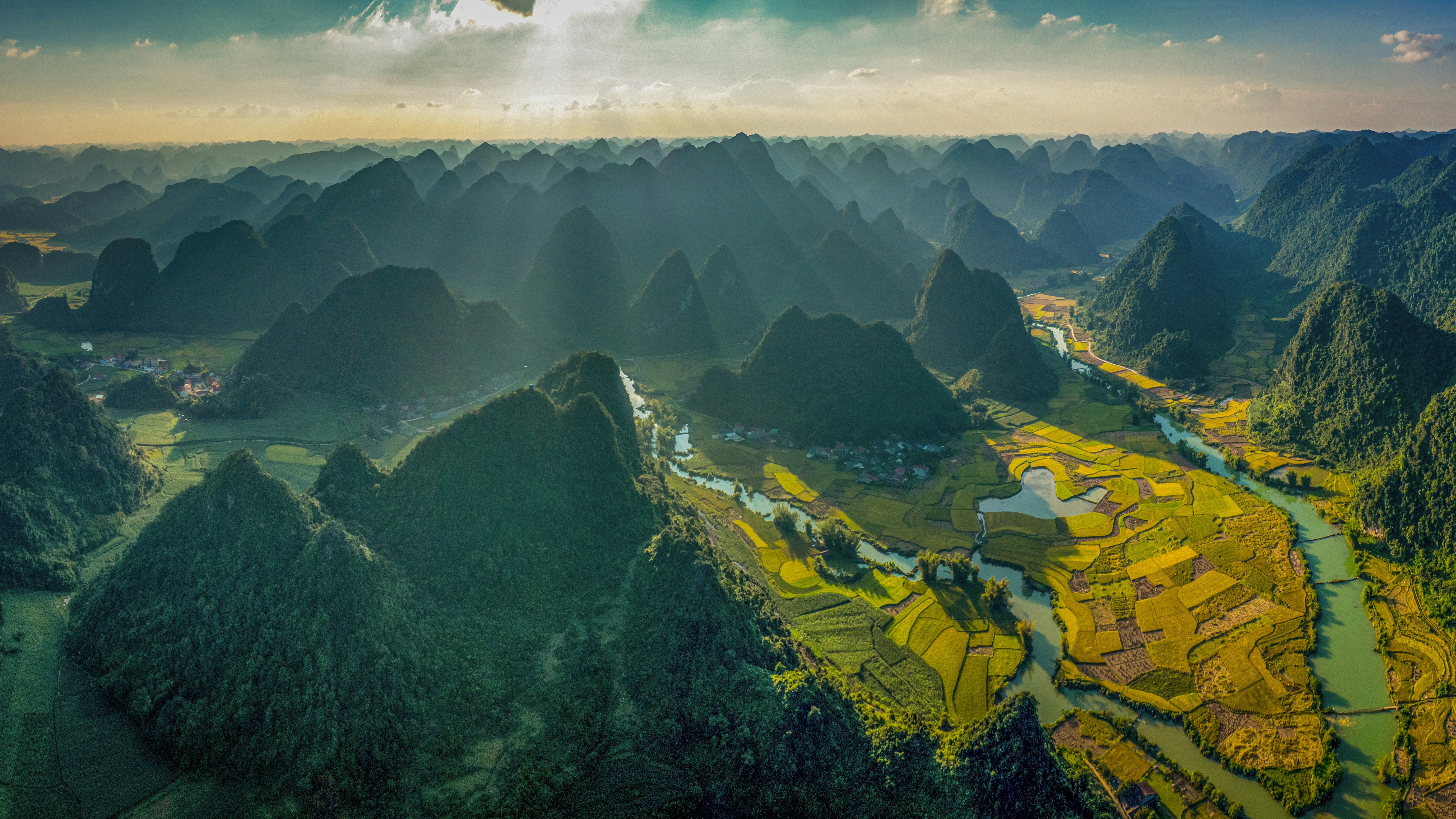 Aerial perspective of rice terraces and winding river amidst lush green hills in Vietnam under a sunlit sky.