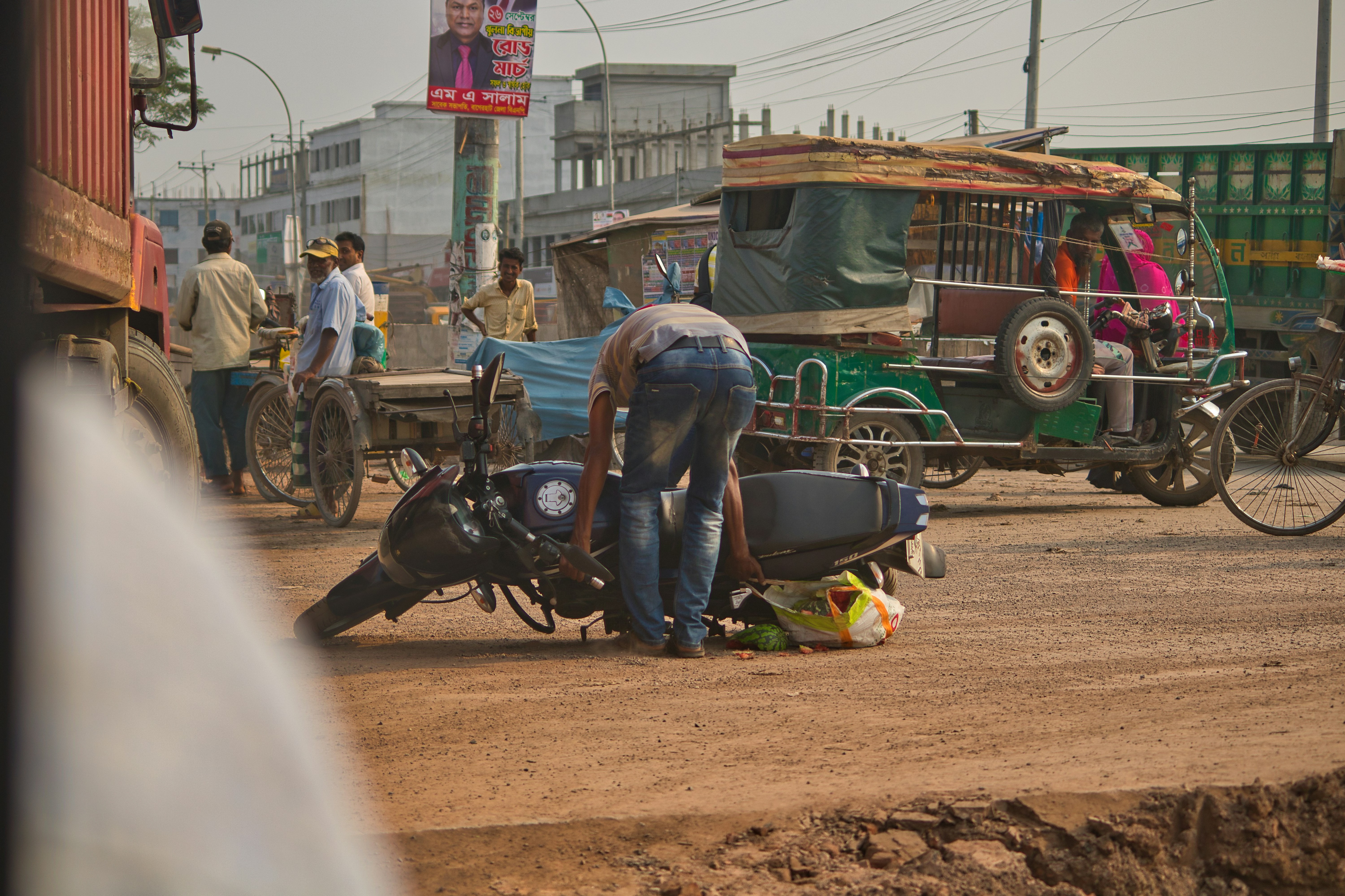 Man in blue shirt and jeans tending to a fallen black motorcycle with a yellow bag on a busy street surrounded by diverse vehicles and cityscape.