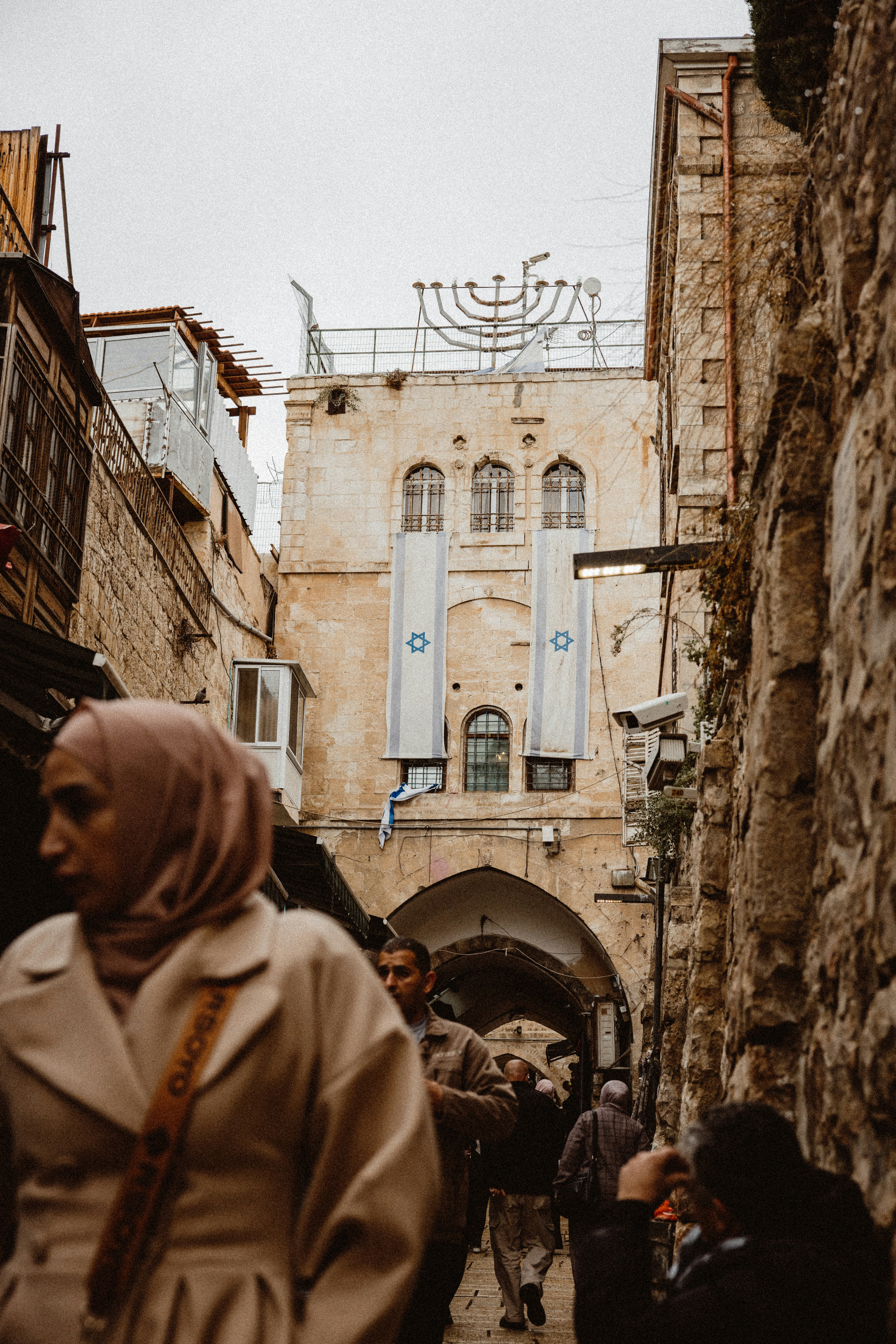 Historic street scene featuring a prominent building adorned with flags and a Menorah, bustling with pedestrians. The atmosphere reflects a blend of tradition and modern life.