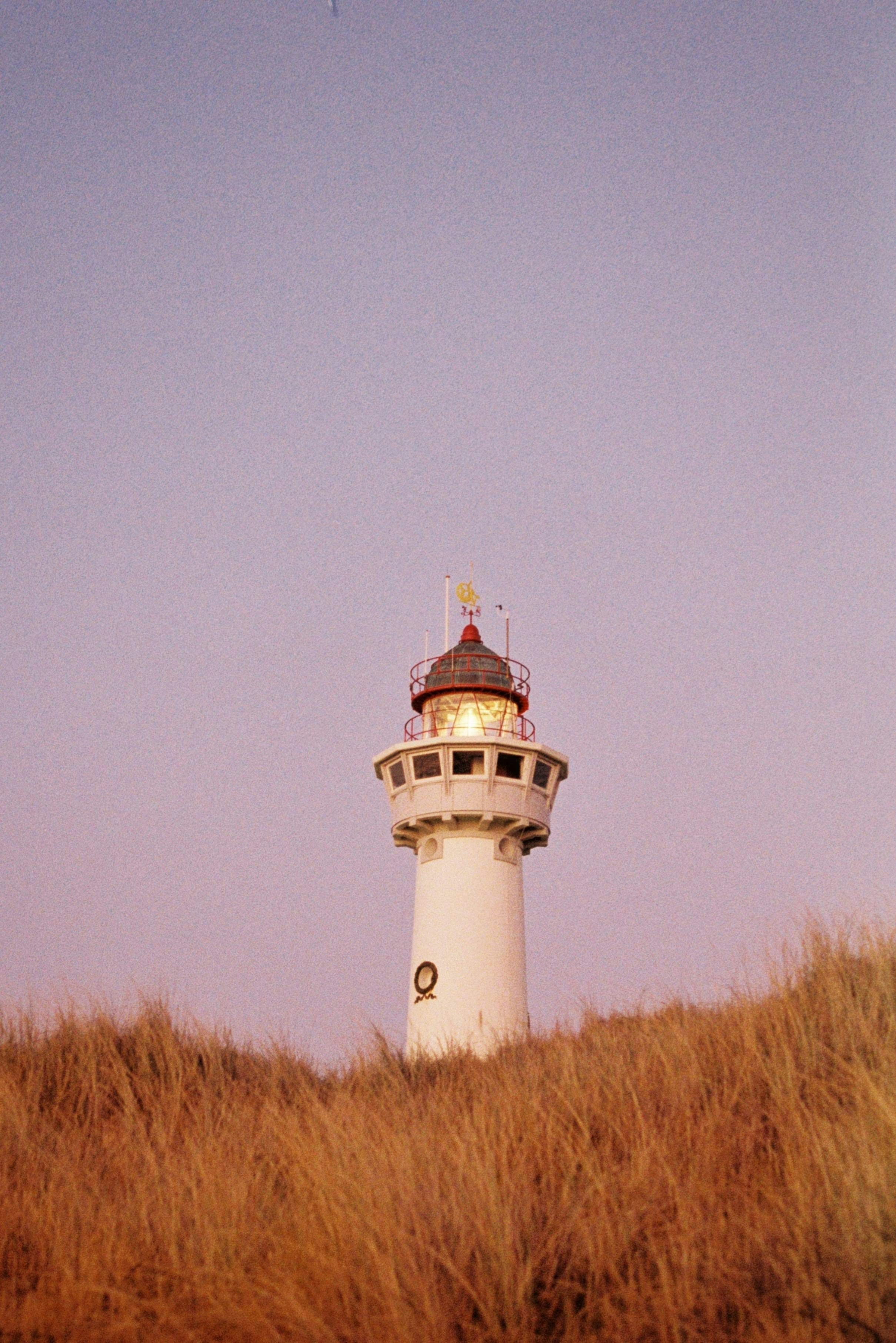 Lighthouse rises above wind-swept dunes with a soft pink-purple sky in the background.