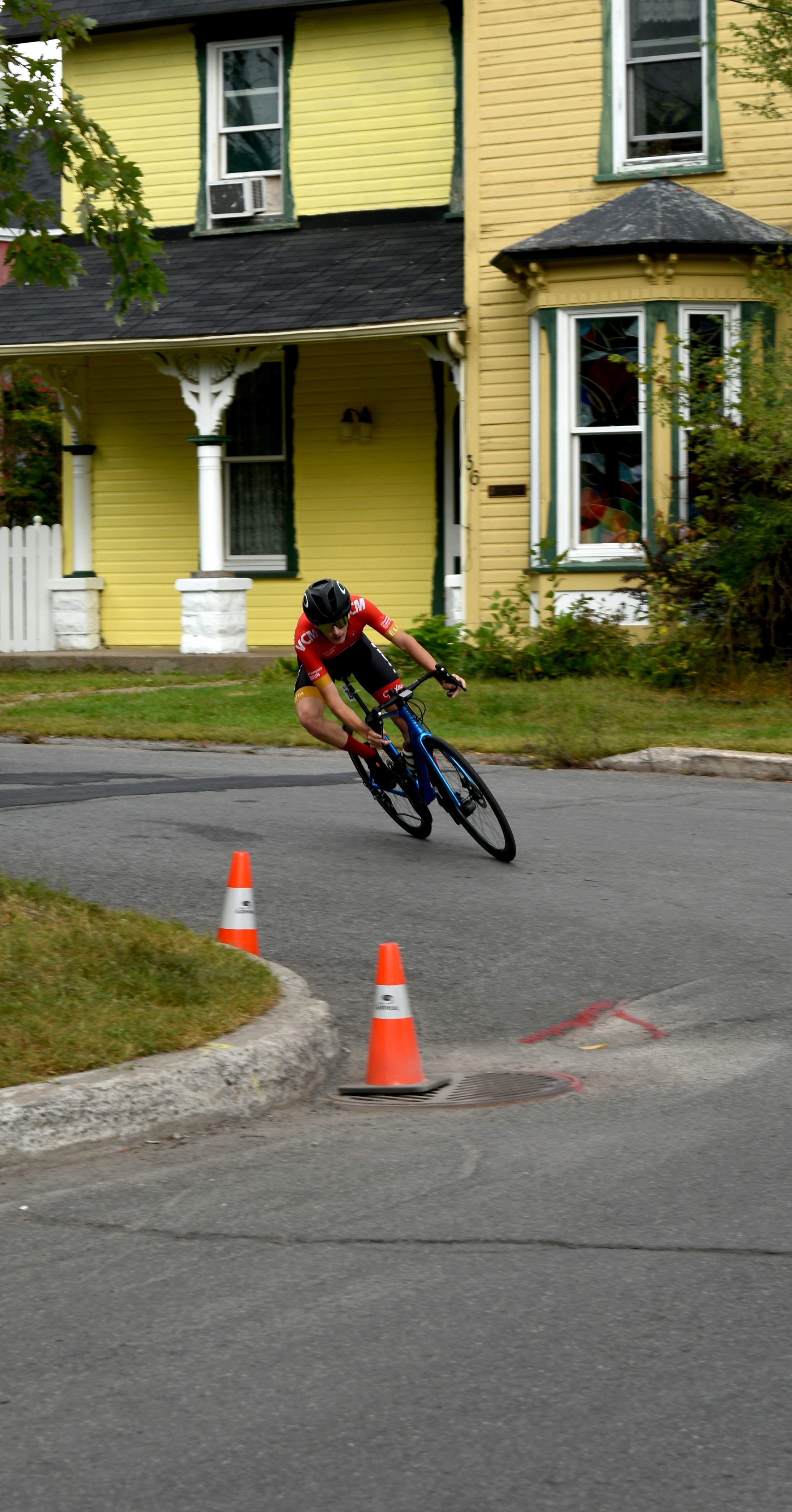 Cyclist navigating a sharp turn on a residential street, with traffic cones marking the route. The vibrant yellow house provides a striking backdrop.