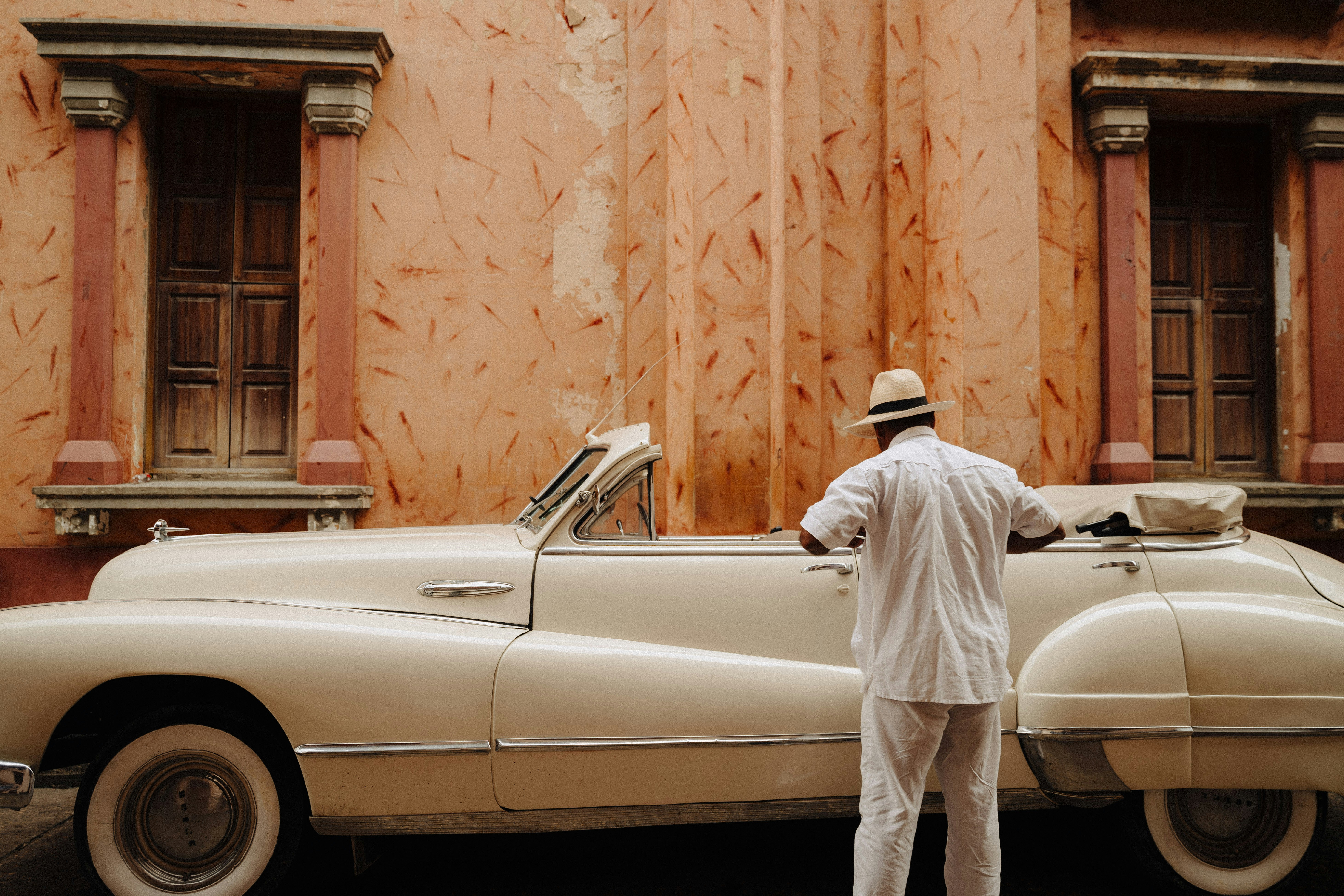 Man in white attire beside a classic cream convertible against a rustic building backdrop.