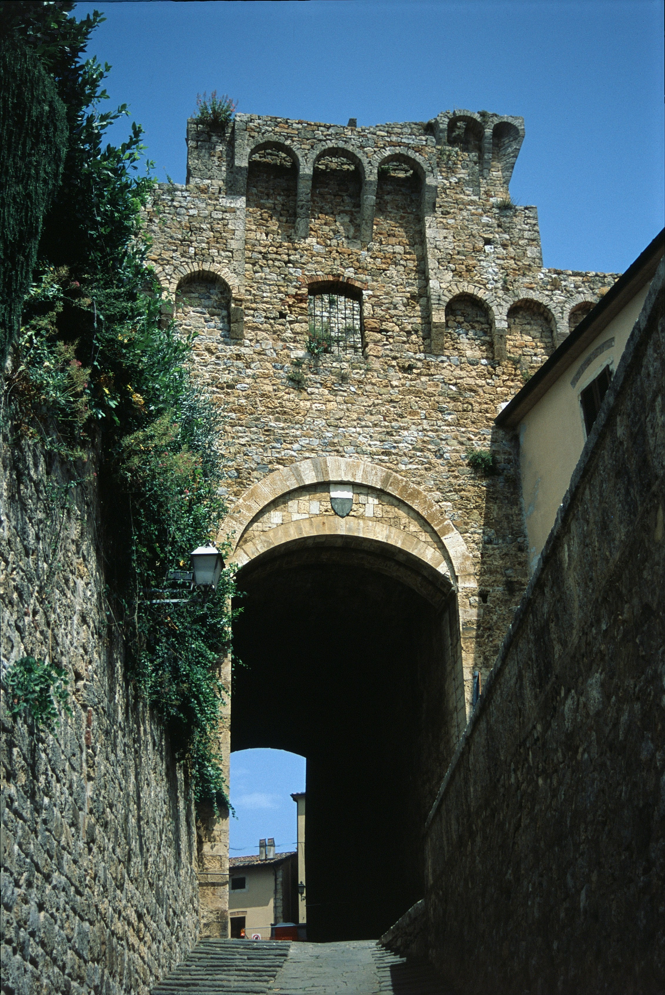 Massive stone fortress arch frames a sunlit stairway into a dark passage. Ivy-clad walls flank the left, with a blue sky above.